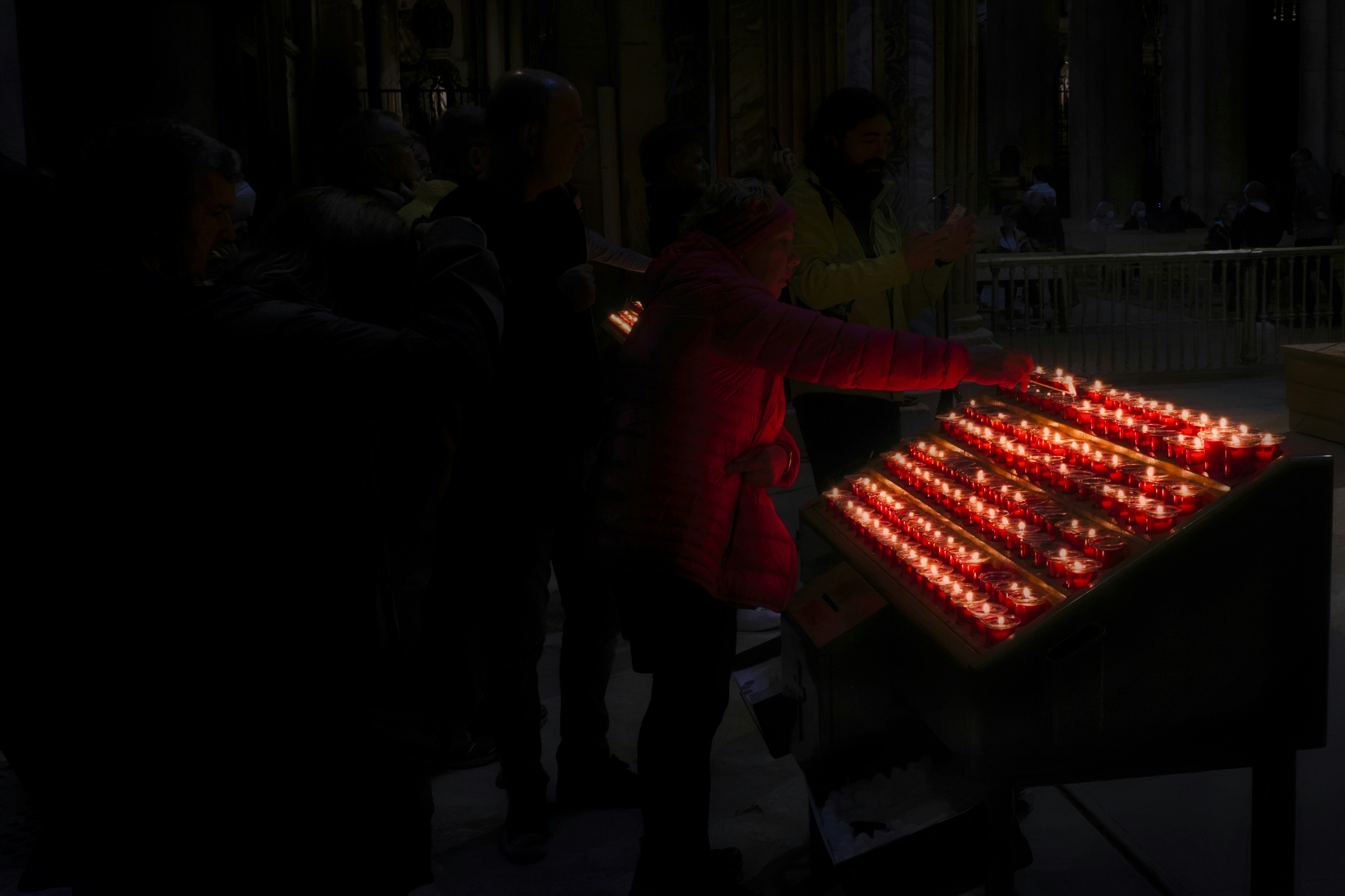 A Person Lighting Candles in a Dark Church Interior - A person in a red jacket reaches out to light a candle on a stand with many burning candles. The candles are lit in rows, creating a warm glow in the otherwise dark interior.