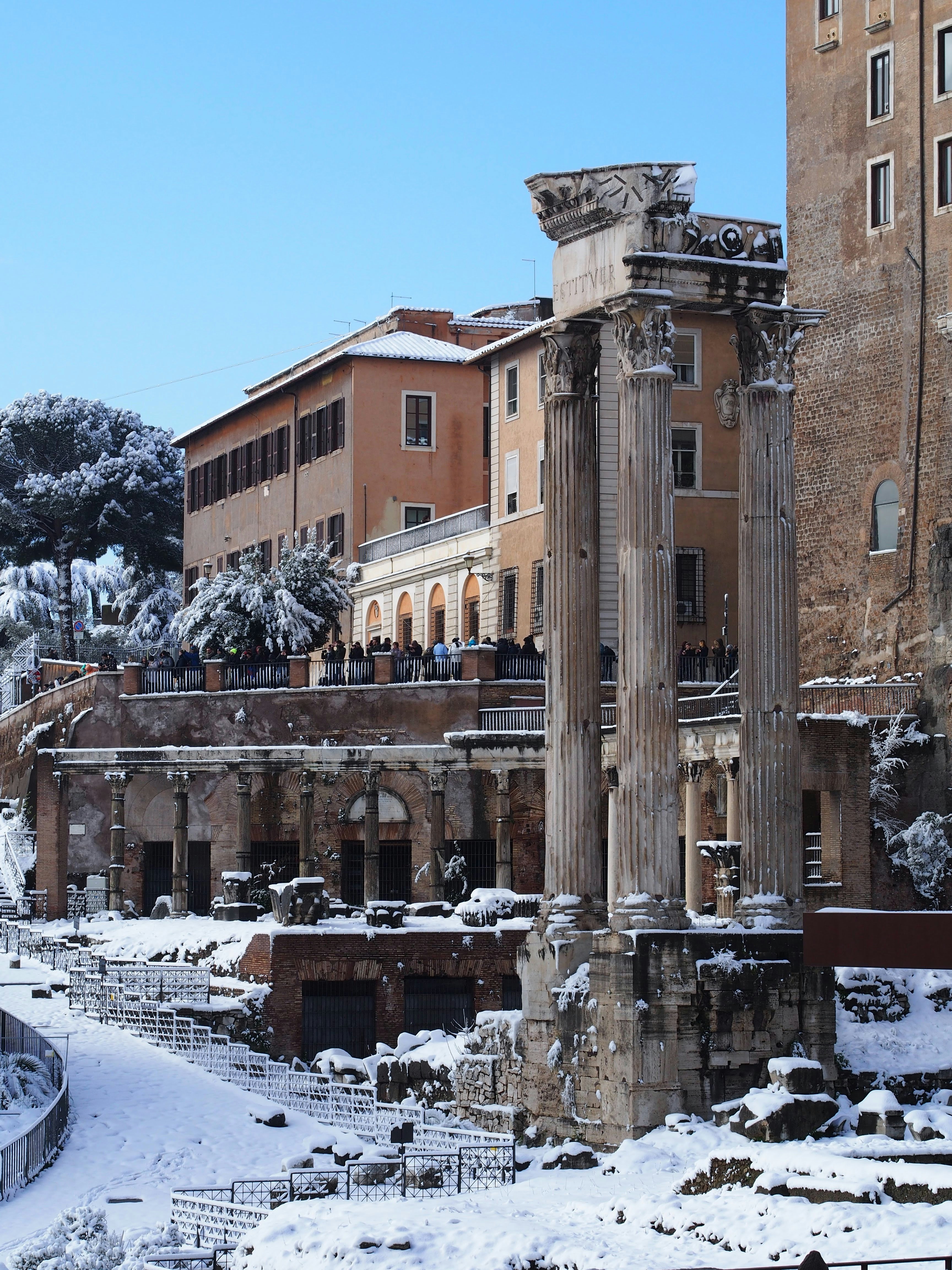Ancient Roman columns stand resilient amidst a blanket of snow, juxtaposed against historic buildings under a clear blue sky.