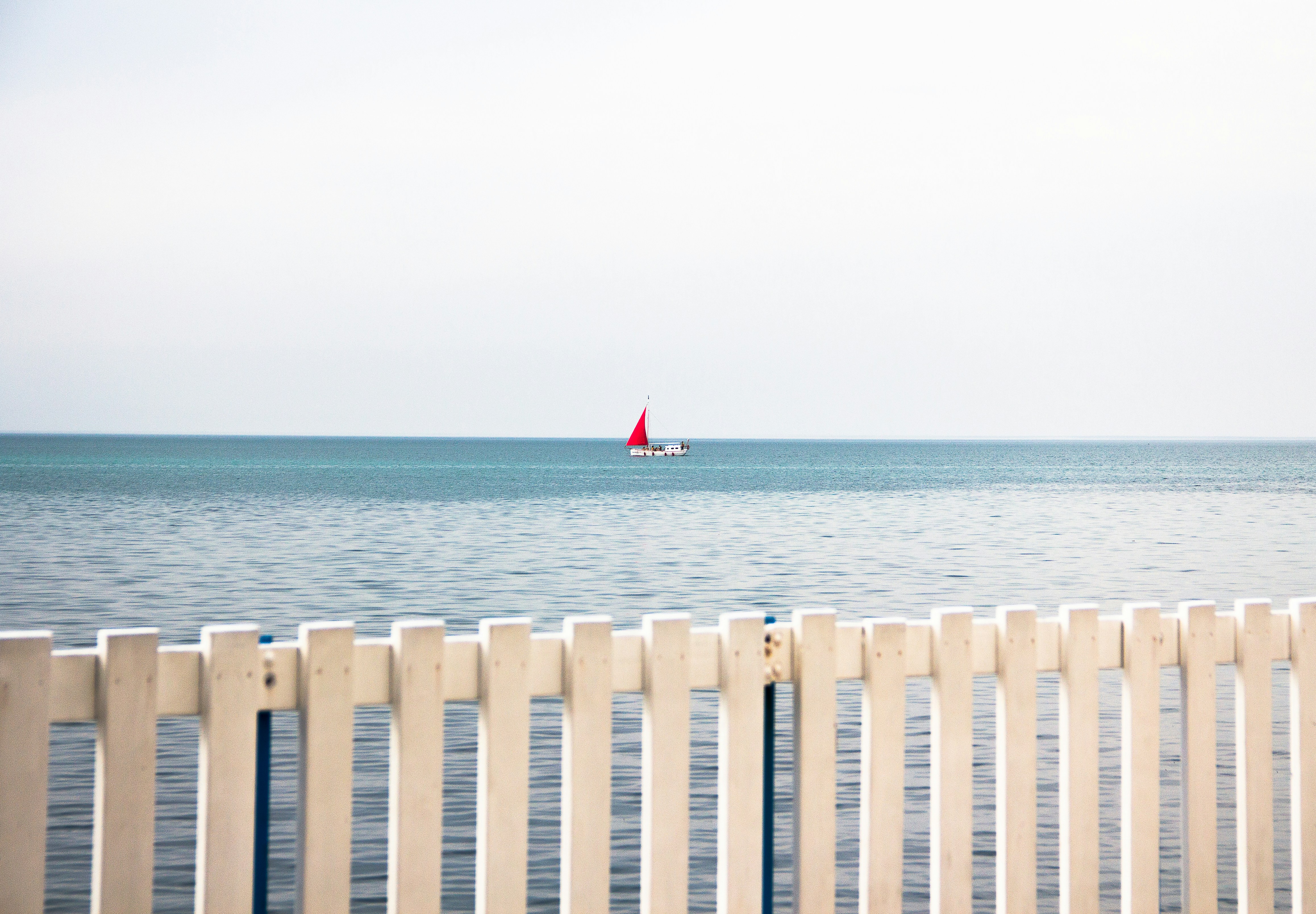 Solitary red sail on a calm horizon behind a pale fence