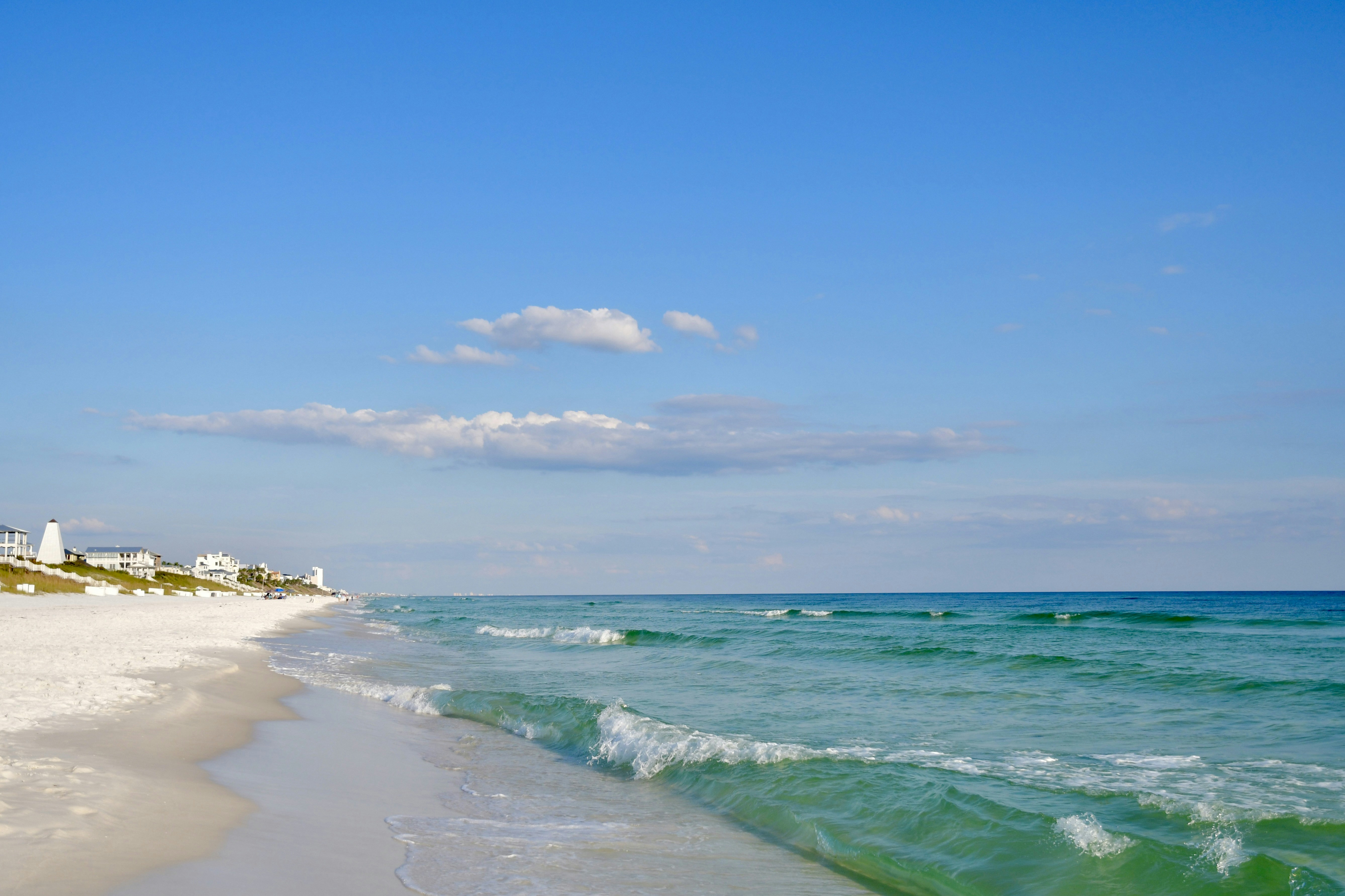 Gentle waves lapping against a pristine sandy beach under a clear blue sky, with distant coastal homes dotting the landscape.