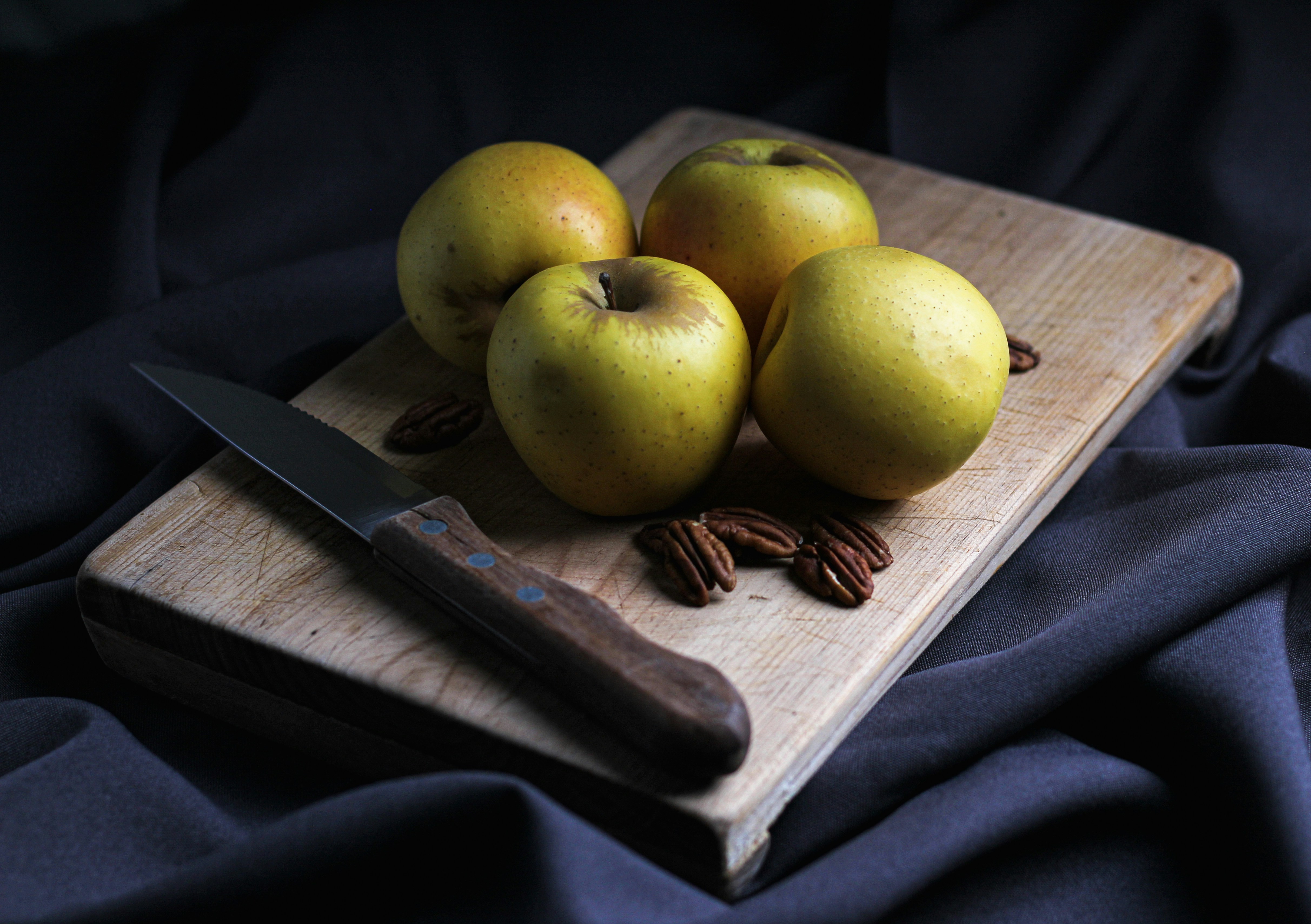 Four golden apples and pecans on a wooden cutting board, accompanied by a knife on a dark fabric backdrop.