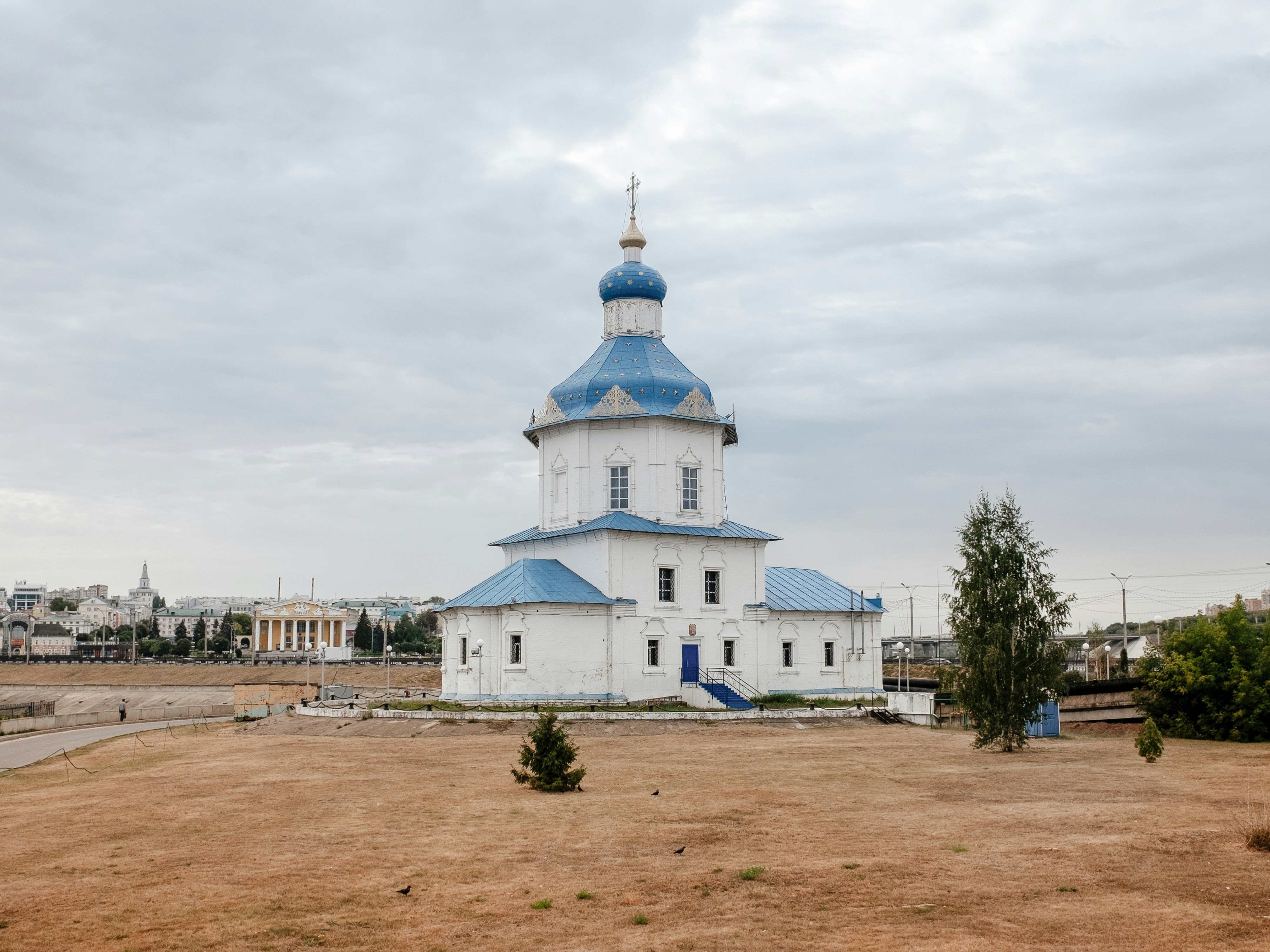 White church with blue domes stands on a dry grassy field under a cloudy sky.