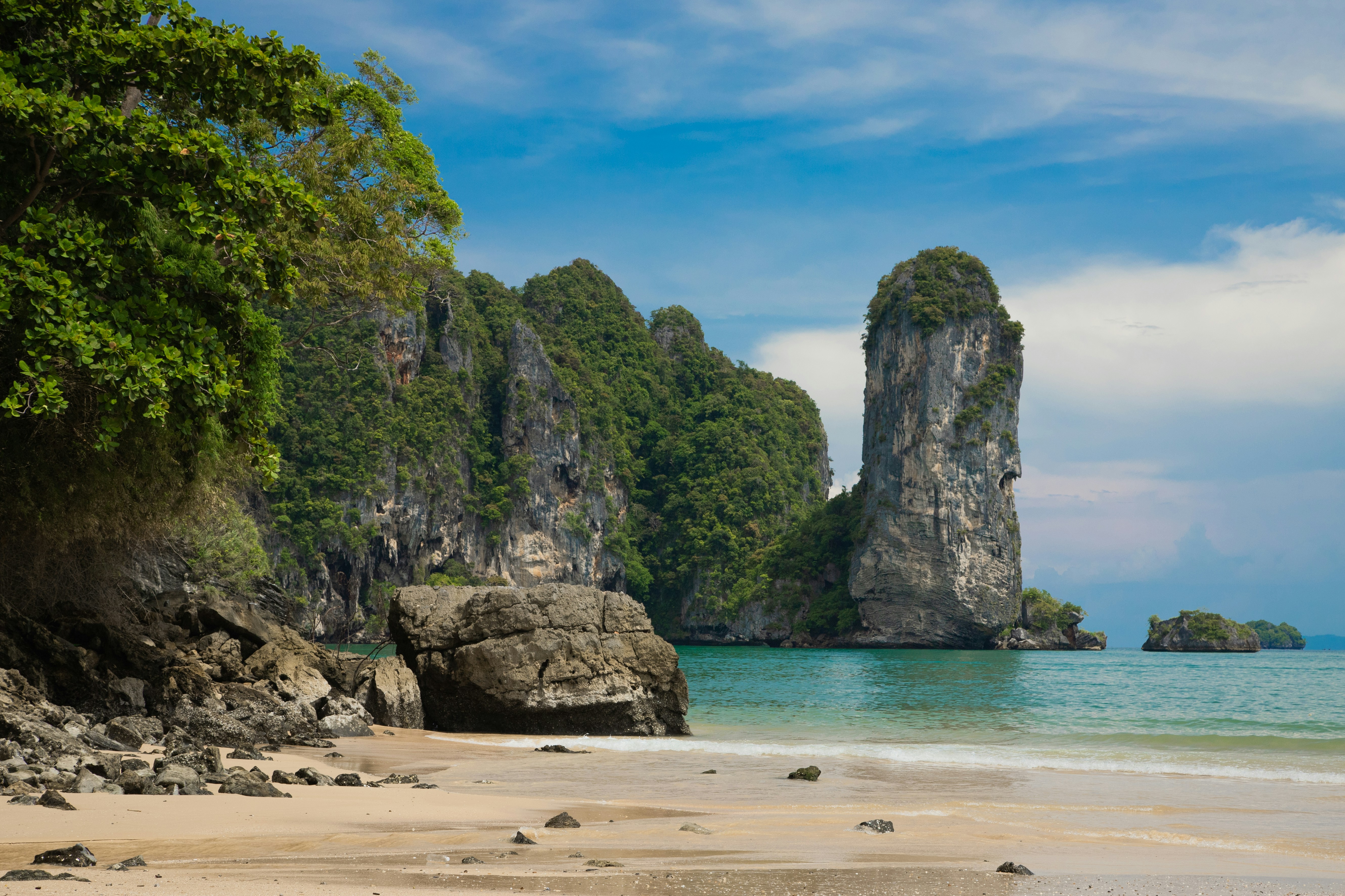 Lush green cliffs rise dramatically from a tranquil beach, with a towering rock formation jutting into the sky against a backdrop of blue skies and gentle waves.