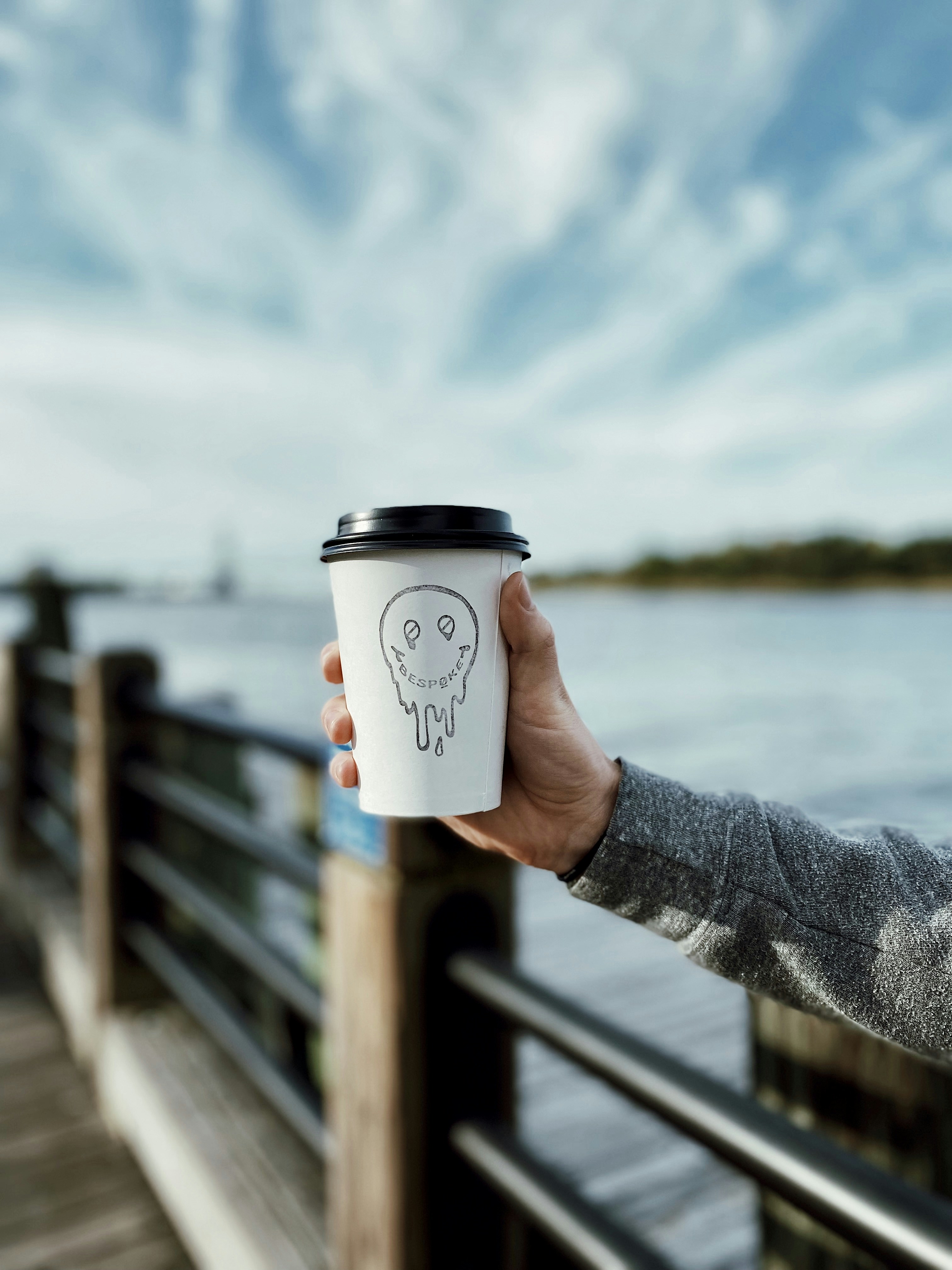 Pierside Sip: Hand-Held Coffee Against Sky and Water