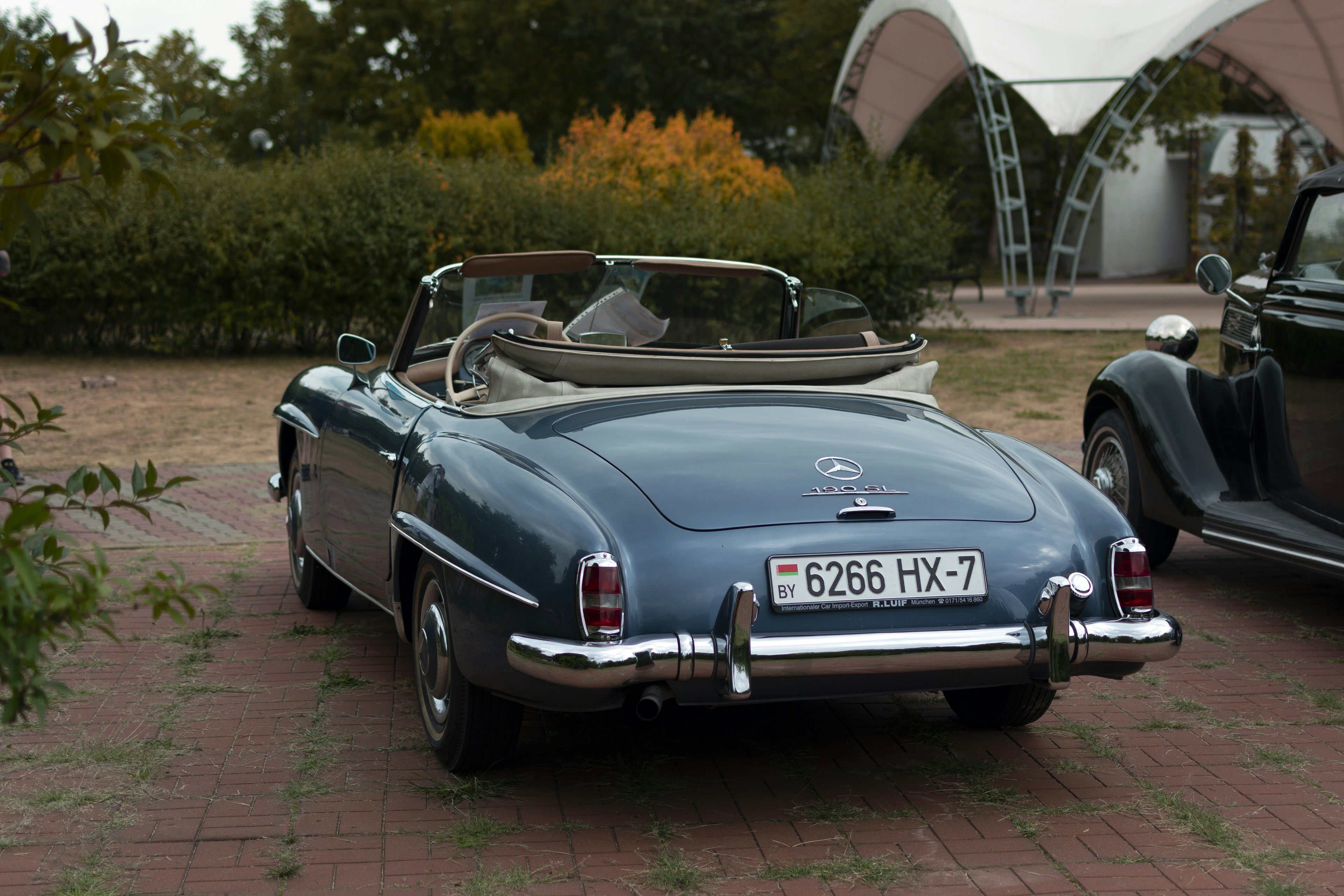 Classic convertible parked on a brick road with lush greenery and a modern arch in the background.