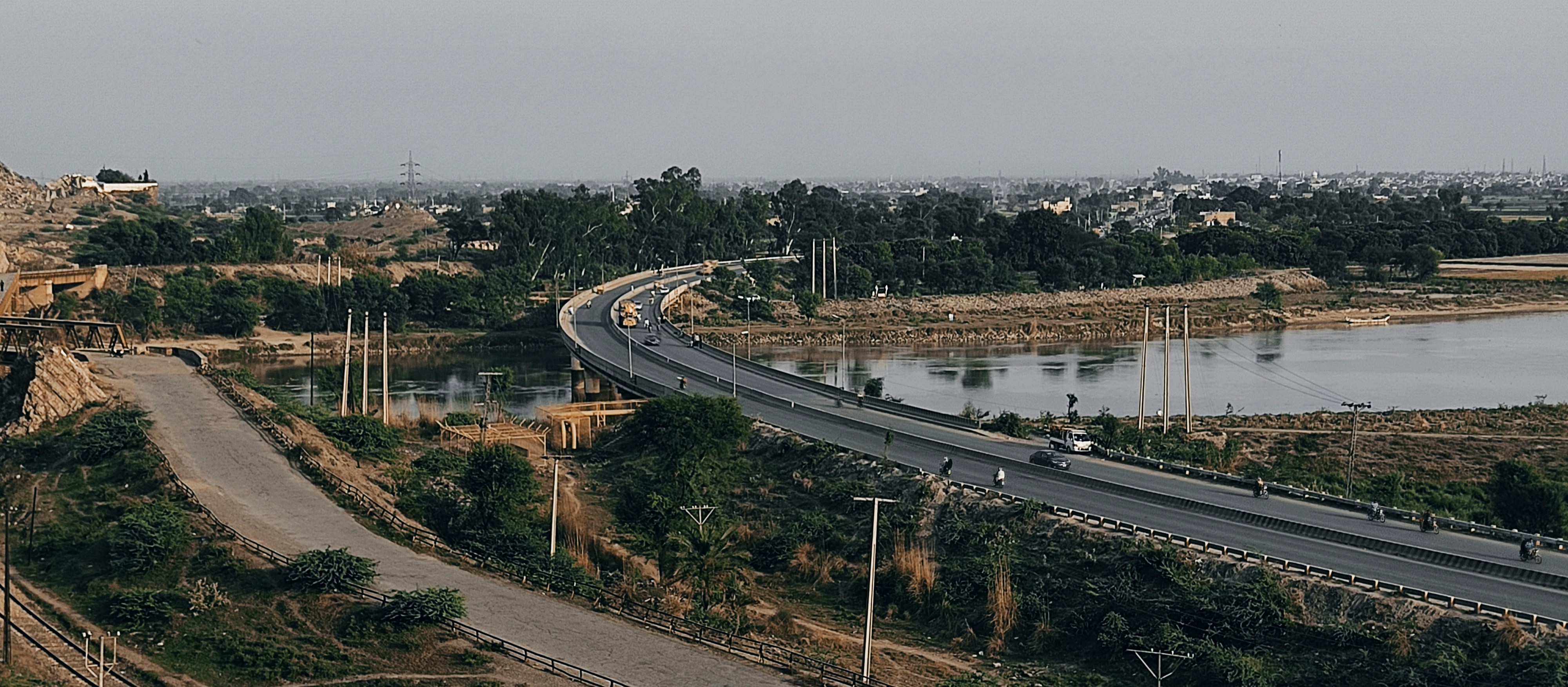 Winding road alongside a tranquil water body, framed by lush greenery and distant cityscape.