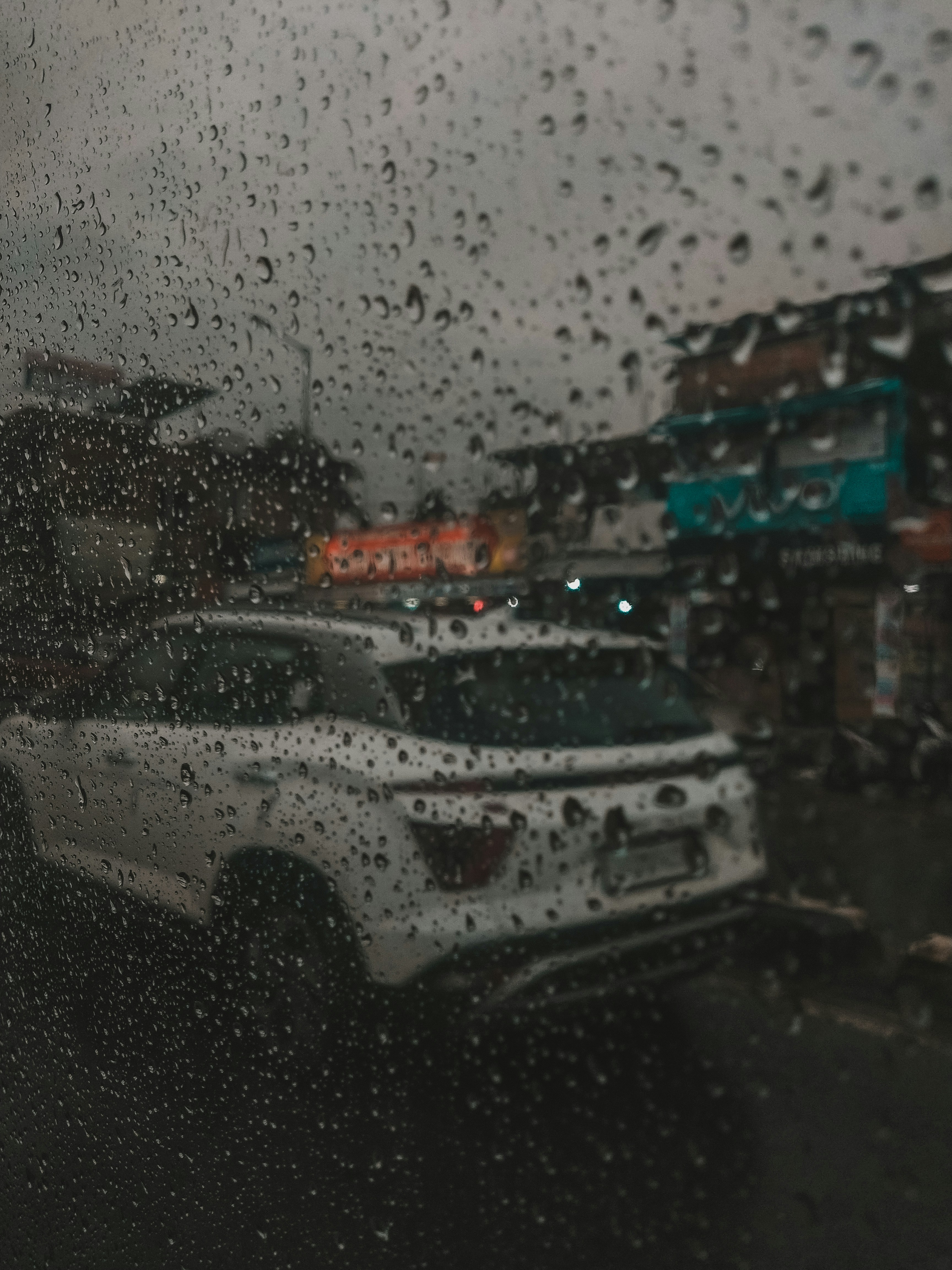 Rain-speckled glass looking out at a blurred urban street with parked cars and storefronts, conveying a moody, rainy-day scene.