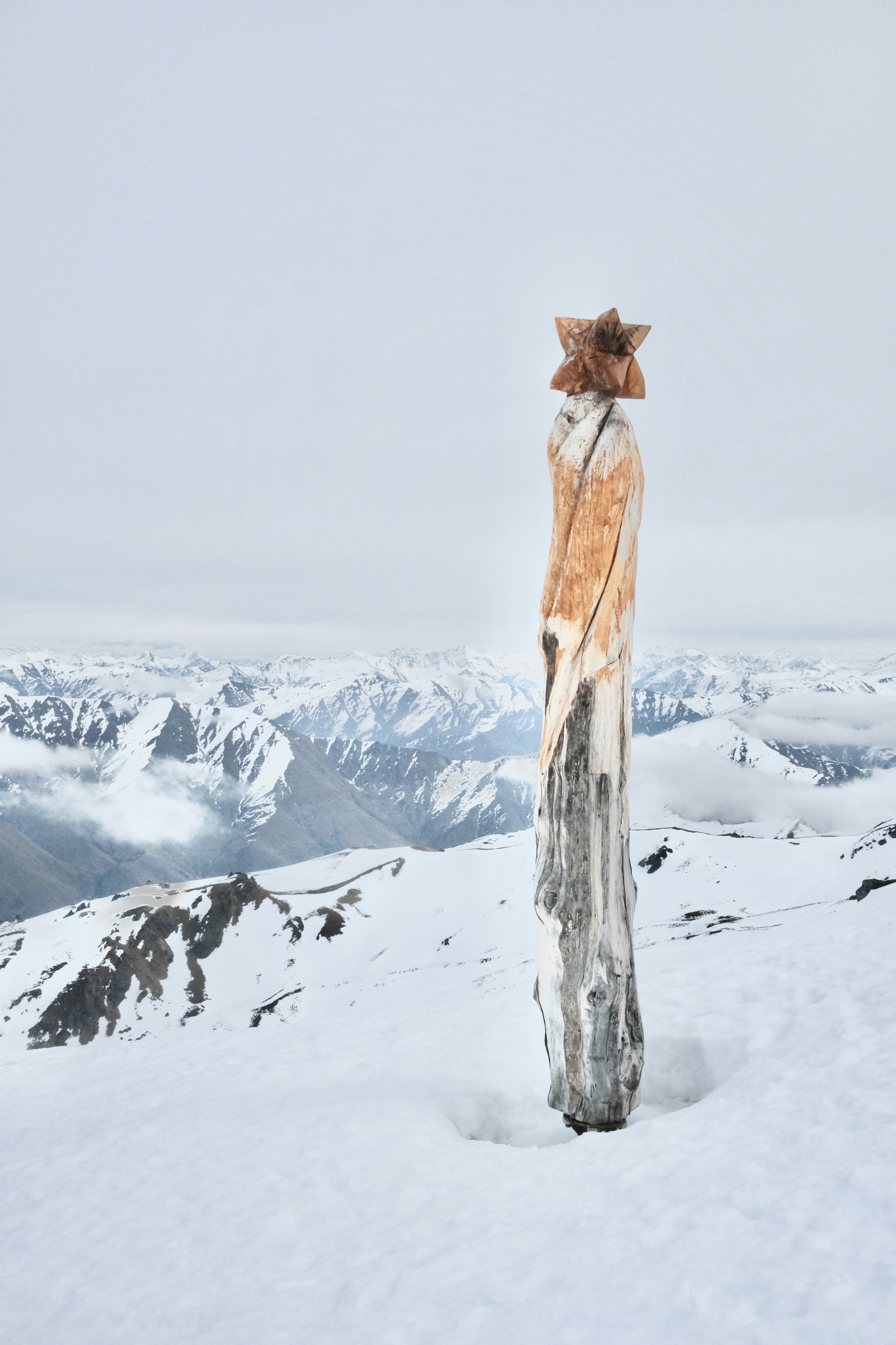 A weathered wooden sculpture stands tall atop a snowy mountain, surrounded by a vast landscape of peaks and clouds.