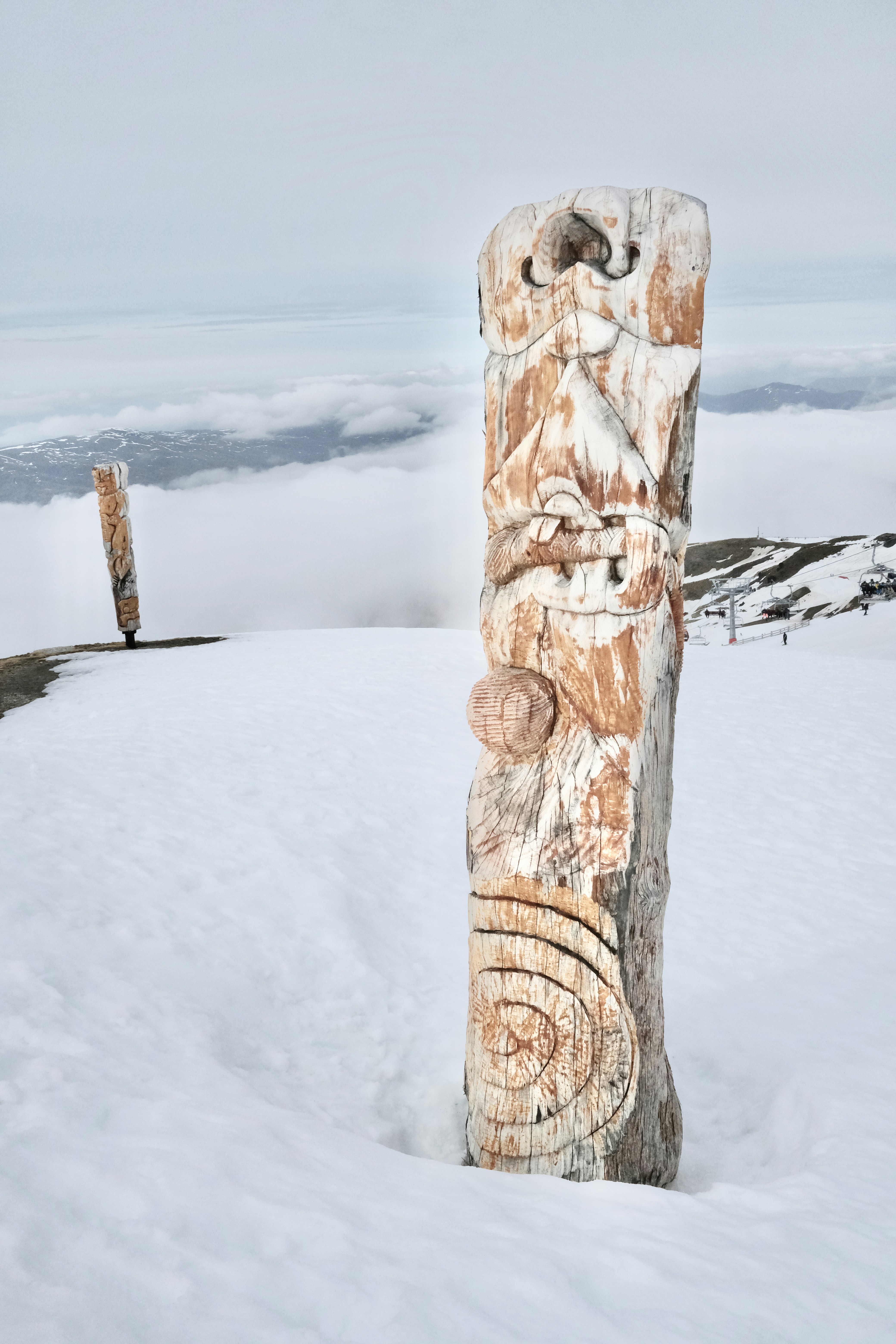 Intricately carved totem stands prominently in a snowy landscape, surrounded by clouds. The wood's texture and patterns tell a story of cultural significance.