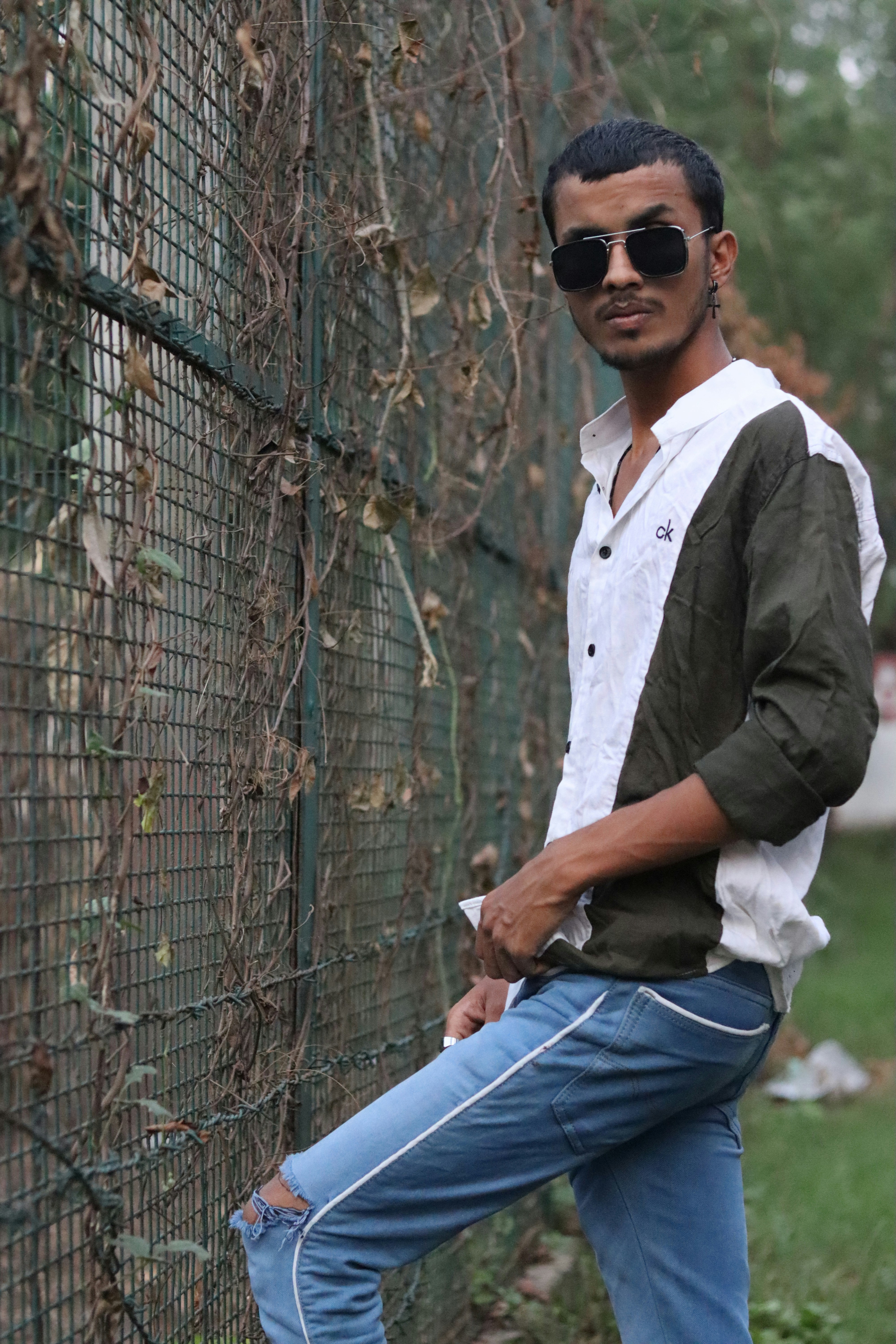 Man in sunglasses posing casually against a wire fence with greenery.