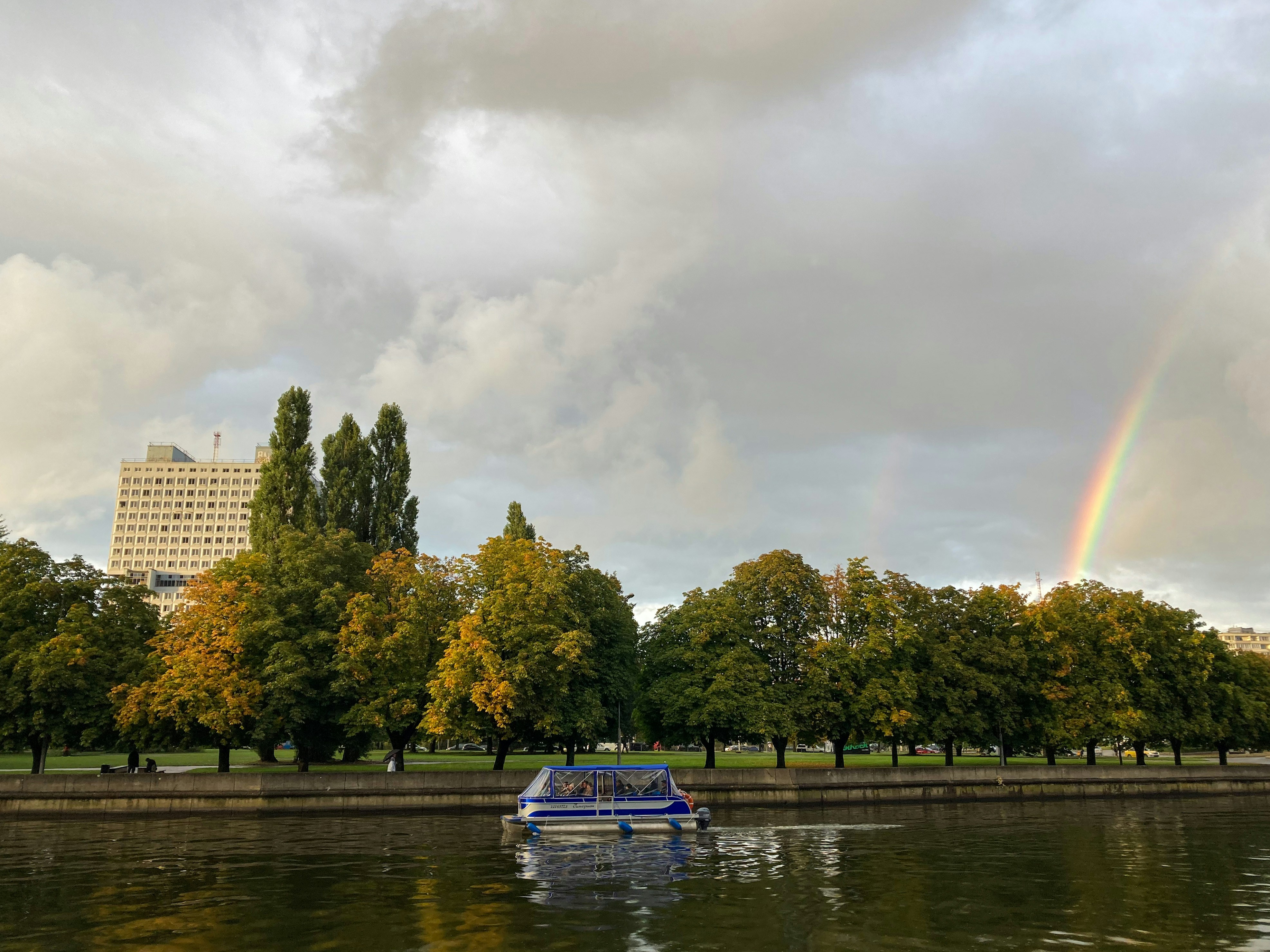 Boat on calm river waters with a line of trees and a vivid rainbow arching against a cloudy sky.