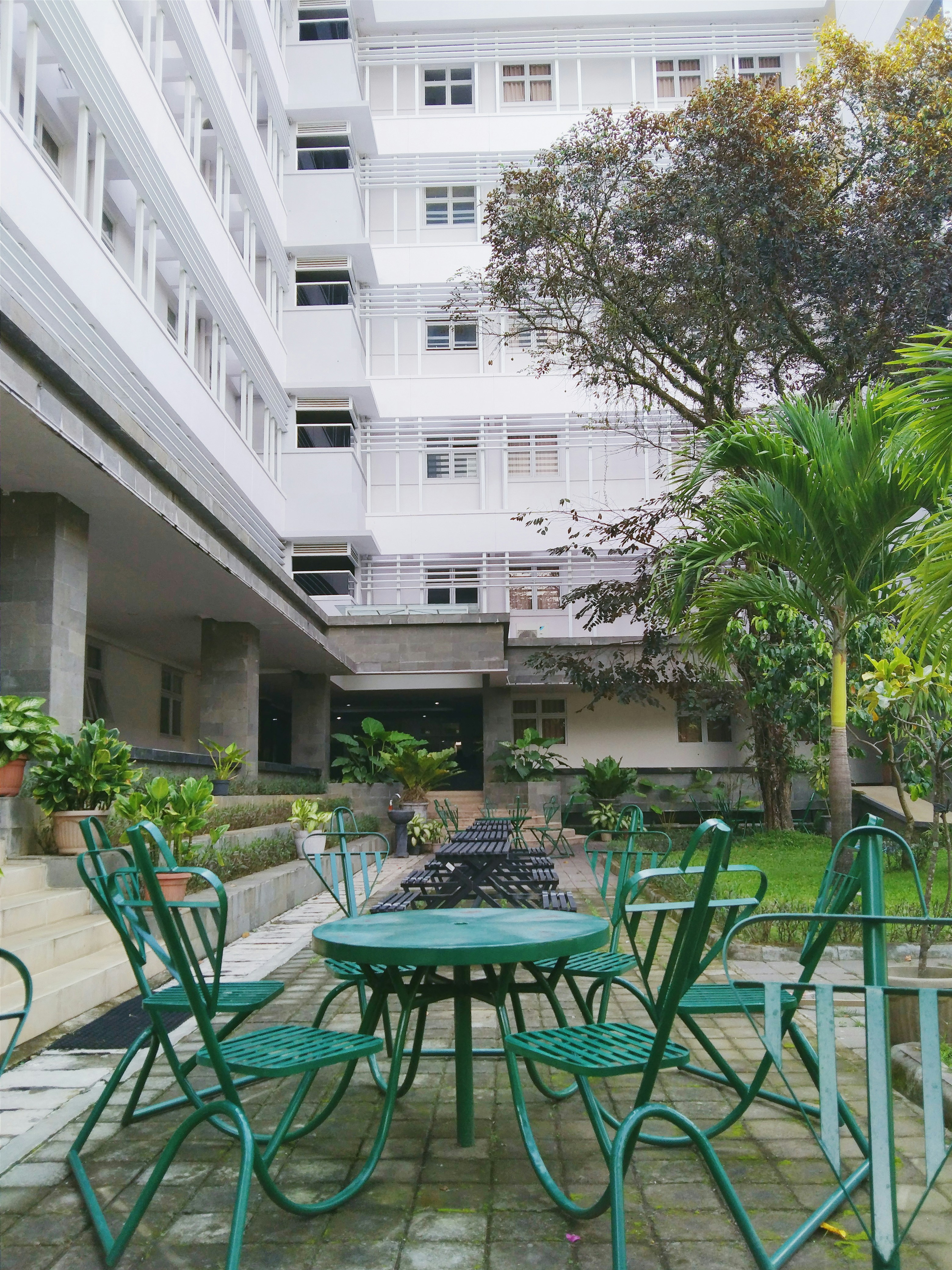Sunlit courtyard between white residential buildings features teal metal chairs arranged around round tables.