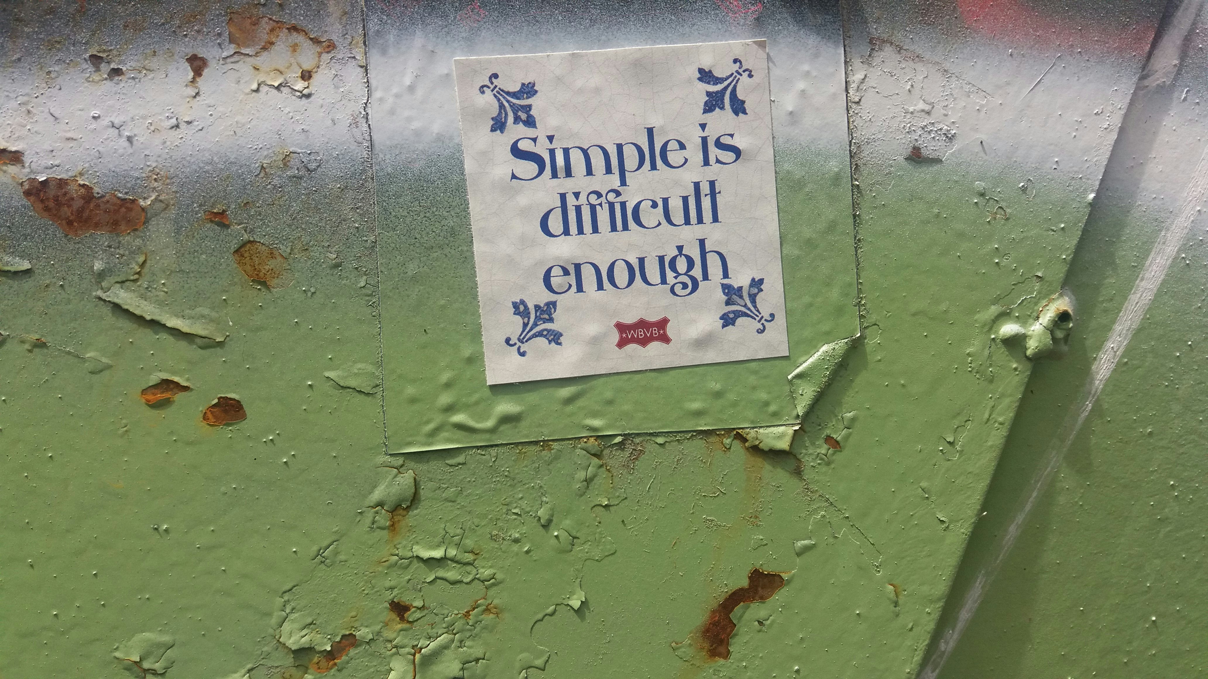 Sticker with the phrase 'Simple is difficult enough' on a chipped, green-painted wall; walls show peeling paint and rusty spots, suggesting an urban, weathered setting.