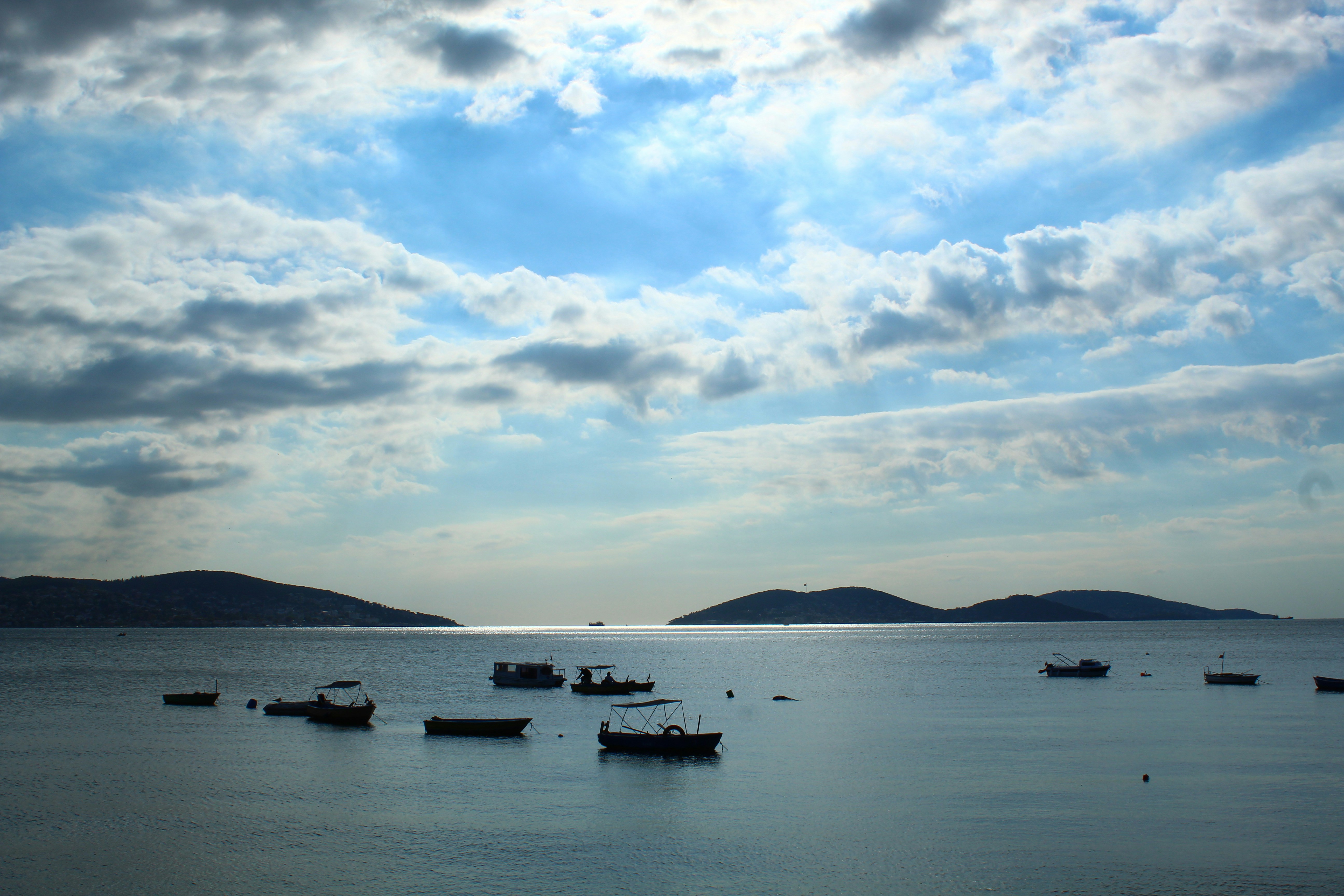 Calm sea with boats silhouetted against a bright sky, islands visible on the horizon.