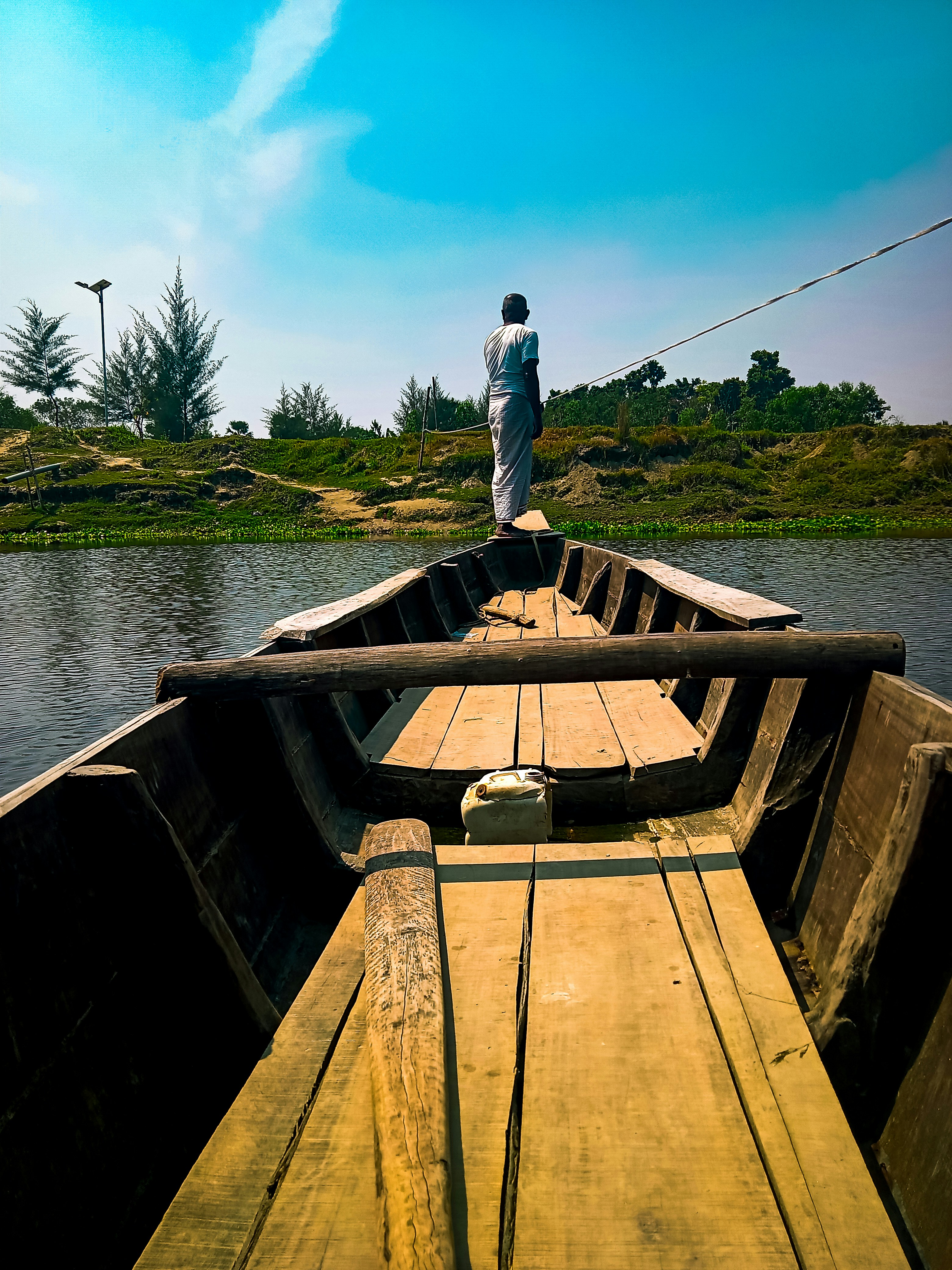 Photograph of a wooden boat on calm water with a lone figure standing at the bow, framed by a bright sky and green shoreline.