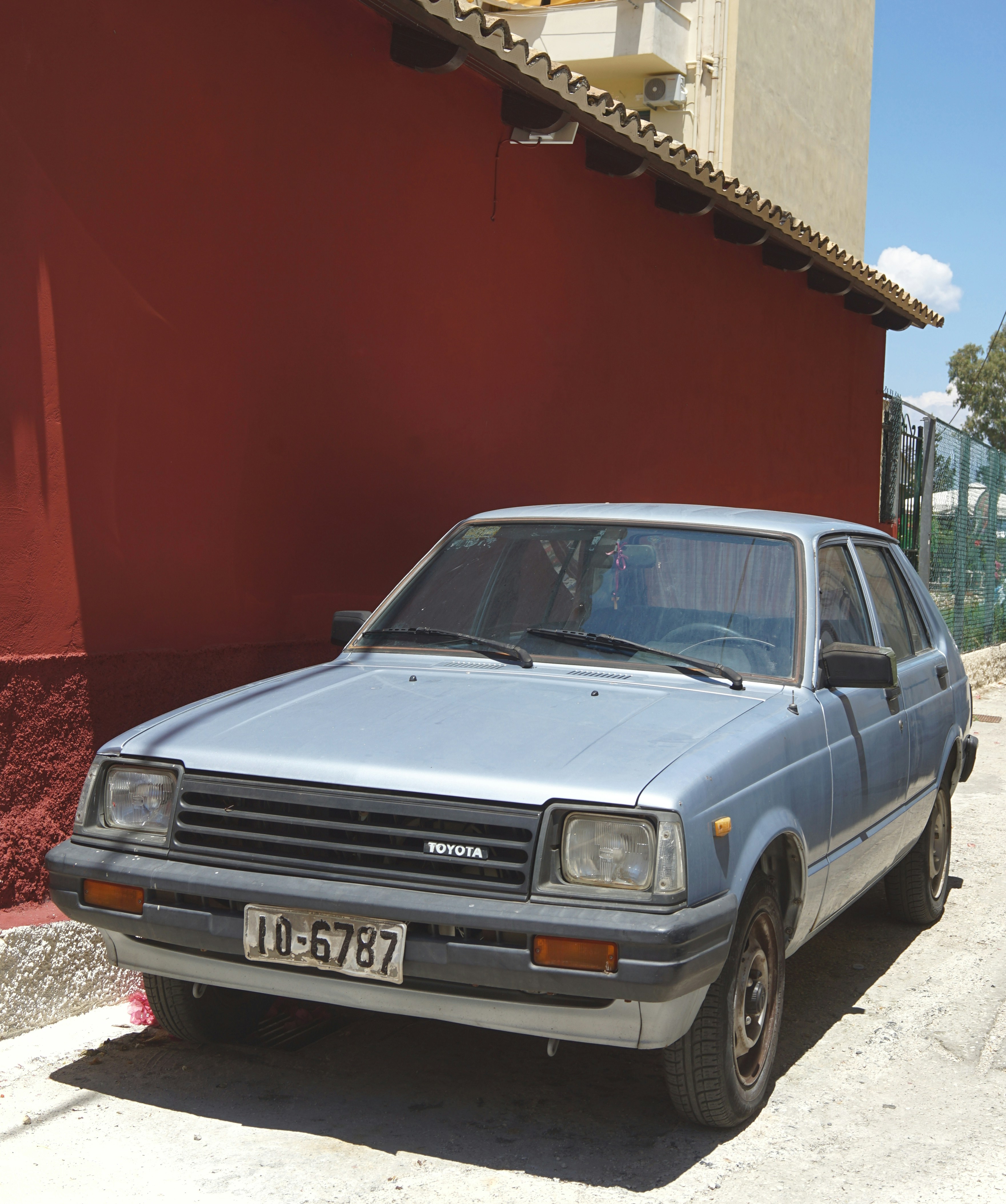 Blue Toyota in a Sunlit Alley Against a Crimson Wall