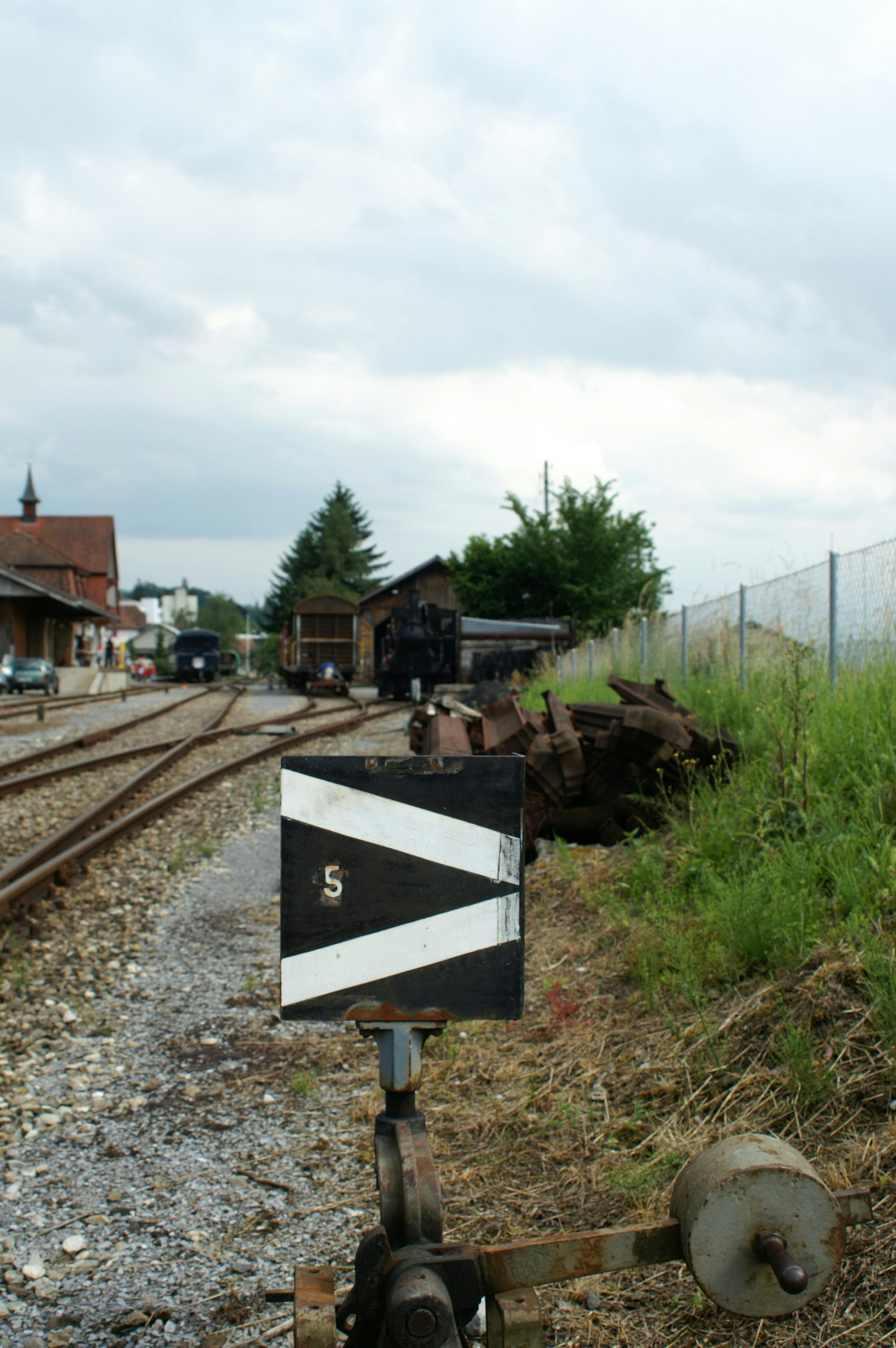 A vintage train signal post stands prominently beside a railway track, with an old station and rolling stock in the background. The scene evokes nostalgia for railway history.