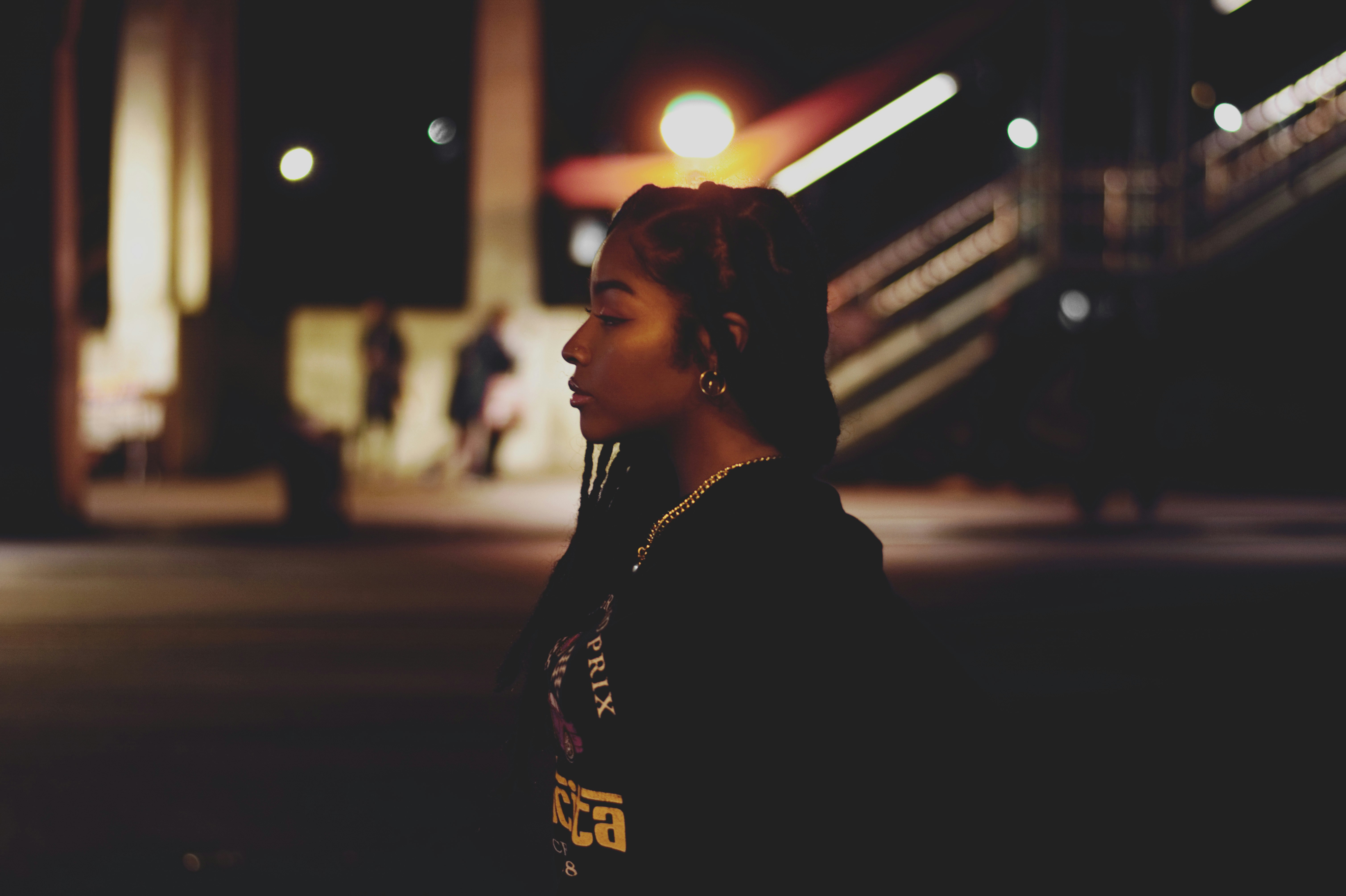 Profile of a young woman against a softly lit urban backdrop, capturing a contemplative moment amidst a bustling scene.