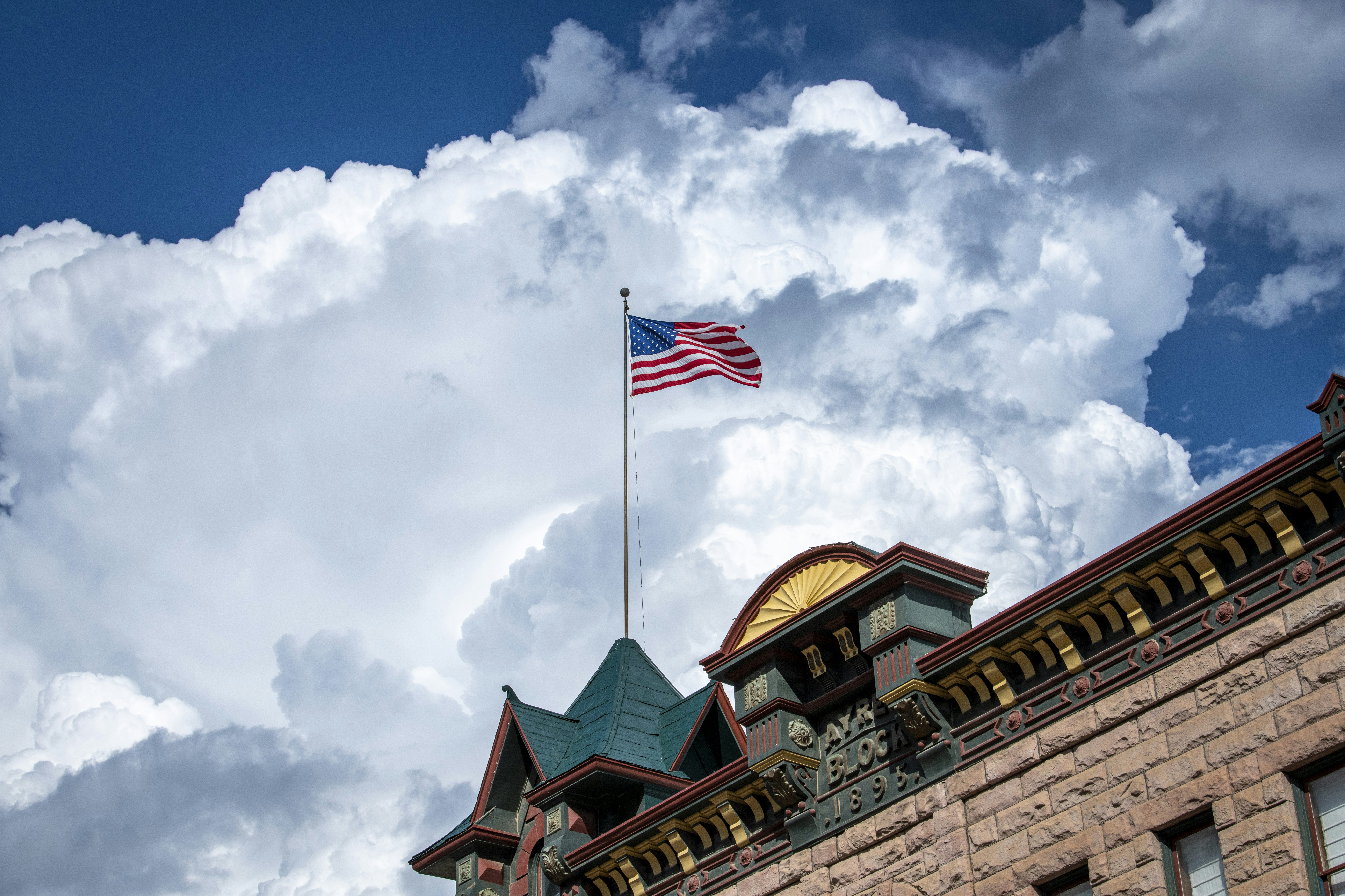 Flag atop a historic brick roofline rises beneath a dramatic cloudscape. Architectural textures along the masonry add depth and contrast to the scene.