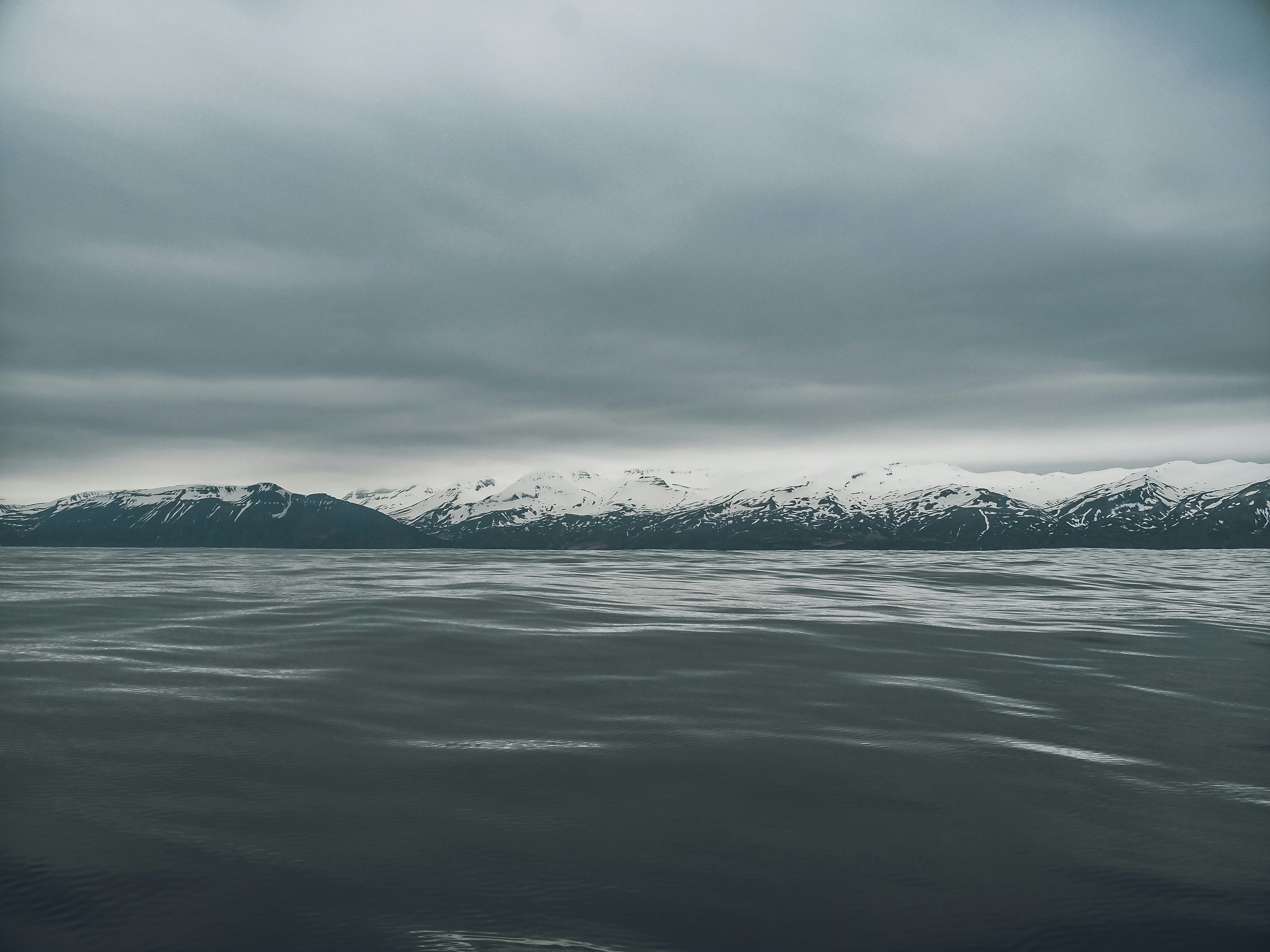 A serene view of a tranquil sea under overcast skies, with snow-capped mountains looming in the distance.