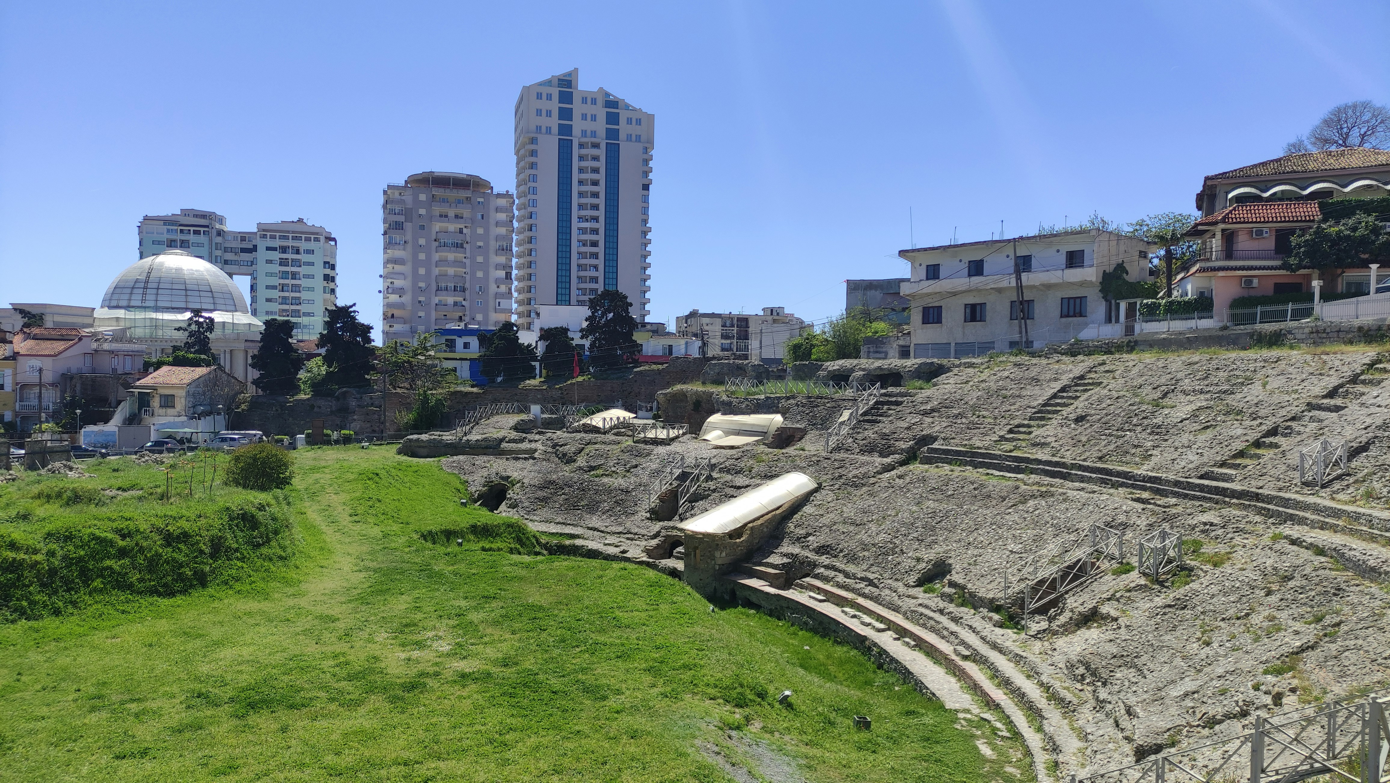 Ancient ruins of a theater juxtaposed with modern city buildings under a clear blue sky.