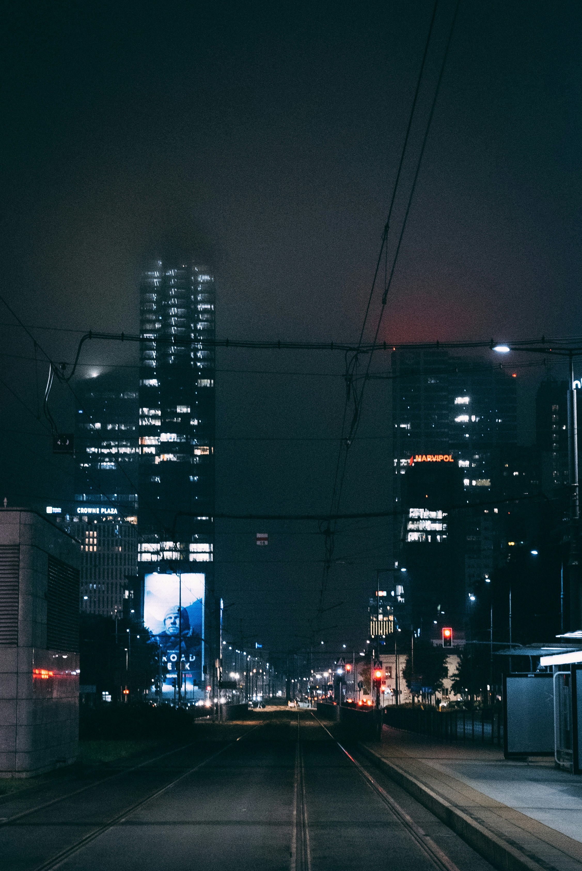 Misty urban street scene illuminated by city lights, featuring towering skyscrapers and a prominent billboard. The atmosphere is dense with fog, creating a mysterious urban landscape.