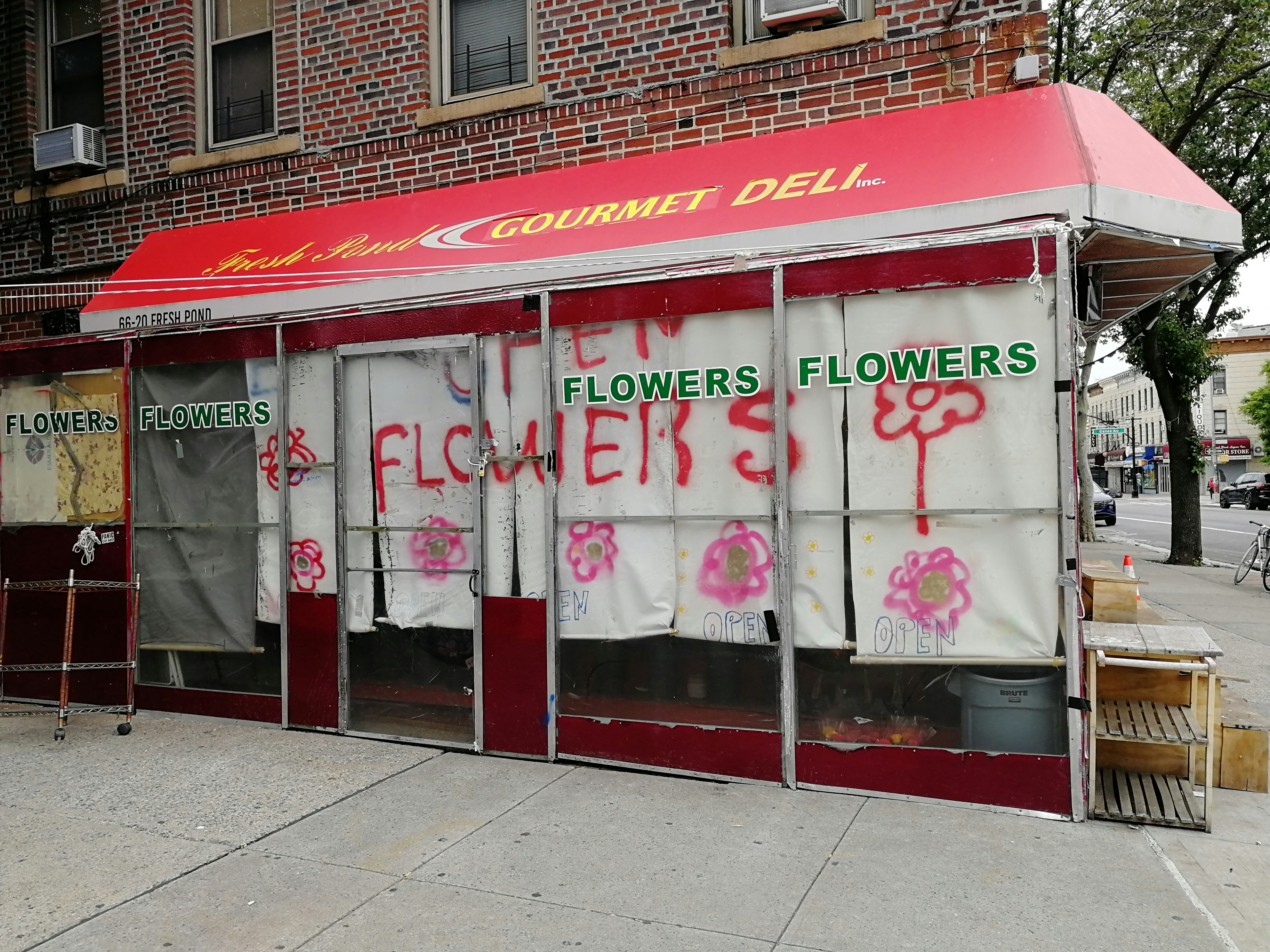 Graffiti-adorned storefront with red awning and 'FLOWERS' signage on translucent panels; urban sidewalk scene highlighting a small deli with a pop of spray-painted art.