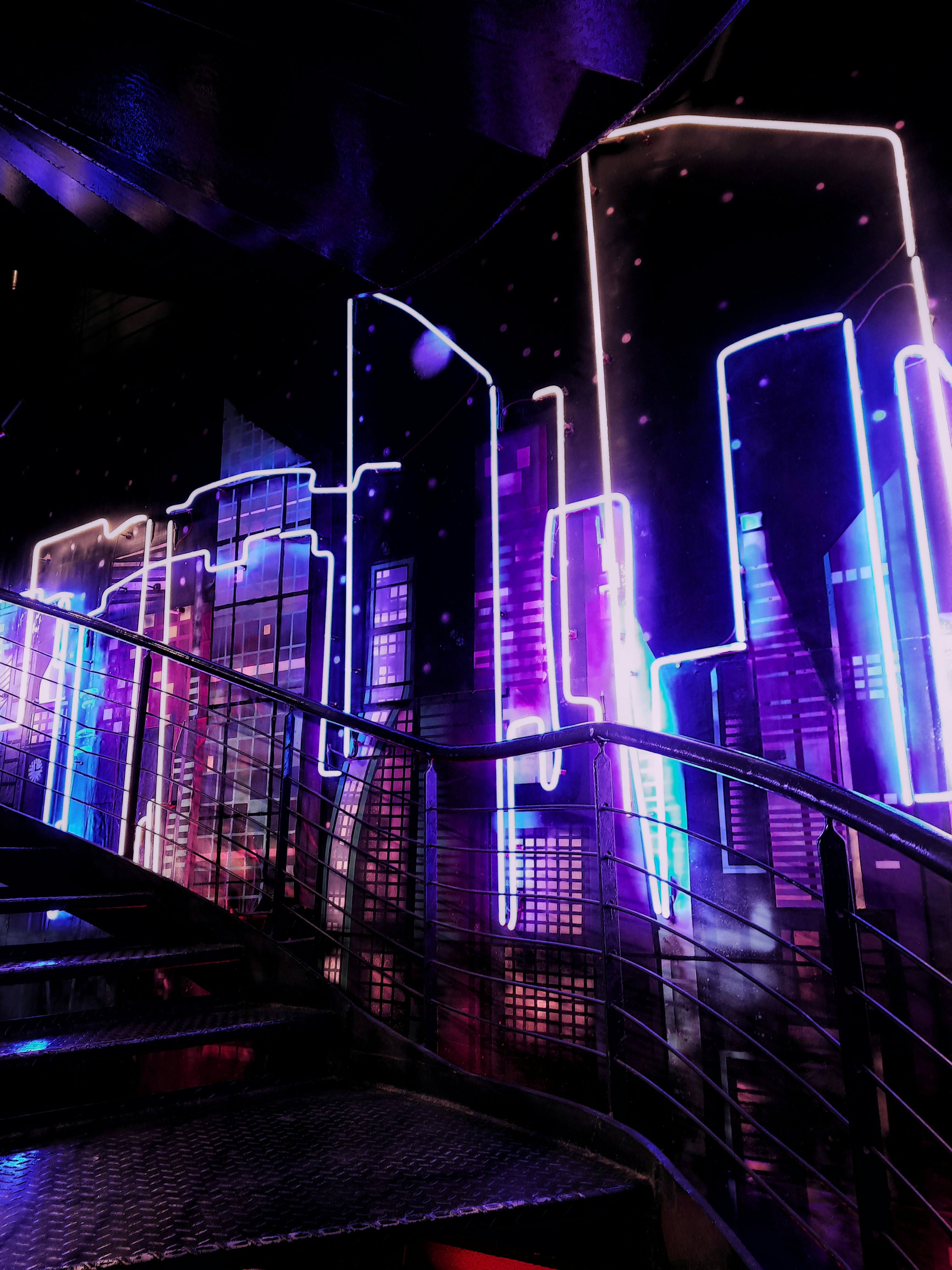 Vibrant neon cityscape mural illuminating a staircase, with a blend of purple and blue tones creating a dynamic atmosphere.