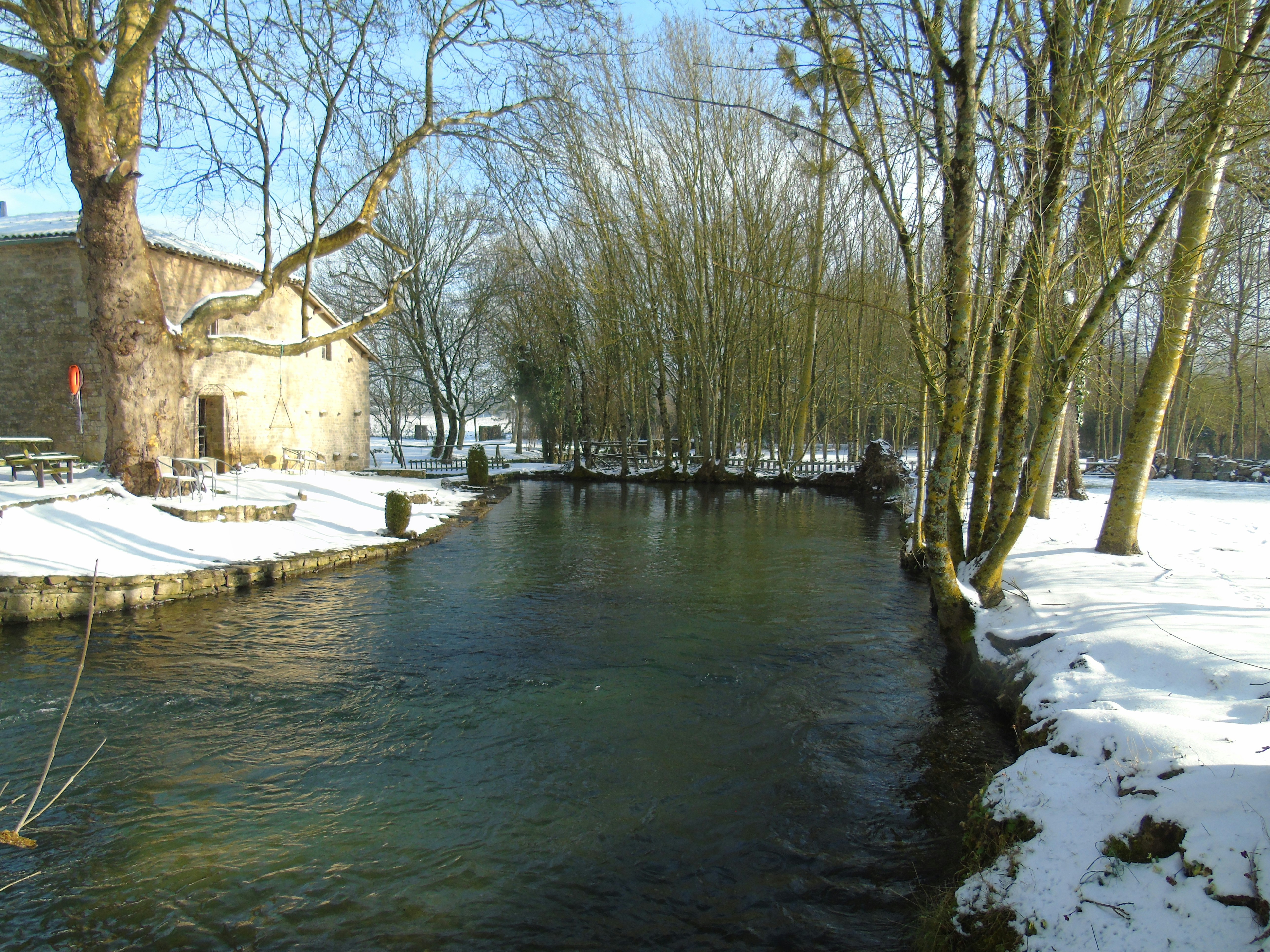 Snow blankets the riverbank as a historic stone mill anchors the left bank. Bare trees line the banks while a small bridge crosses the tranquil water.