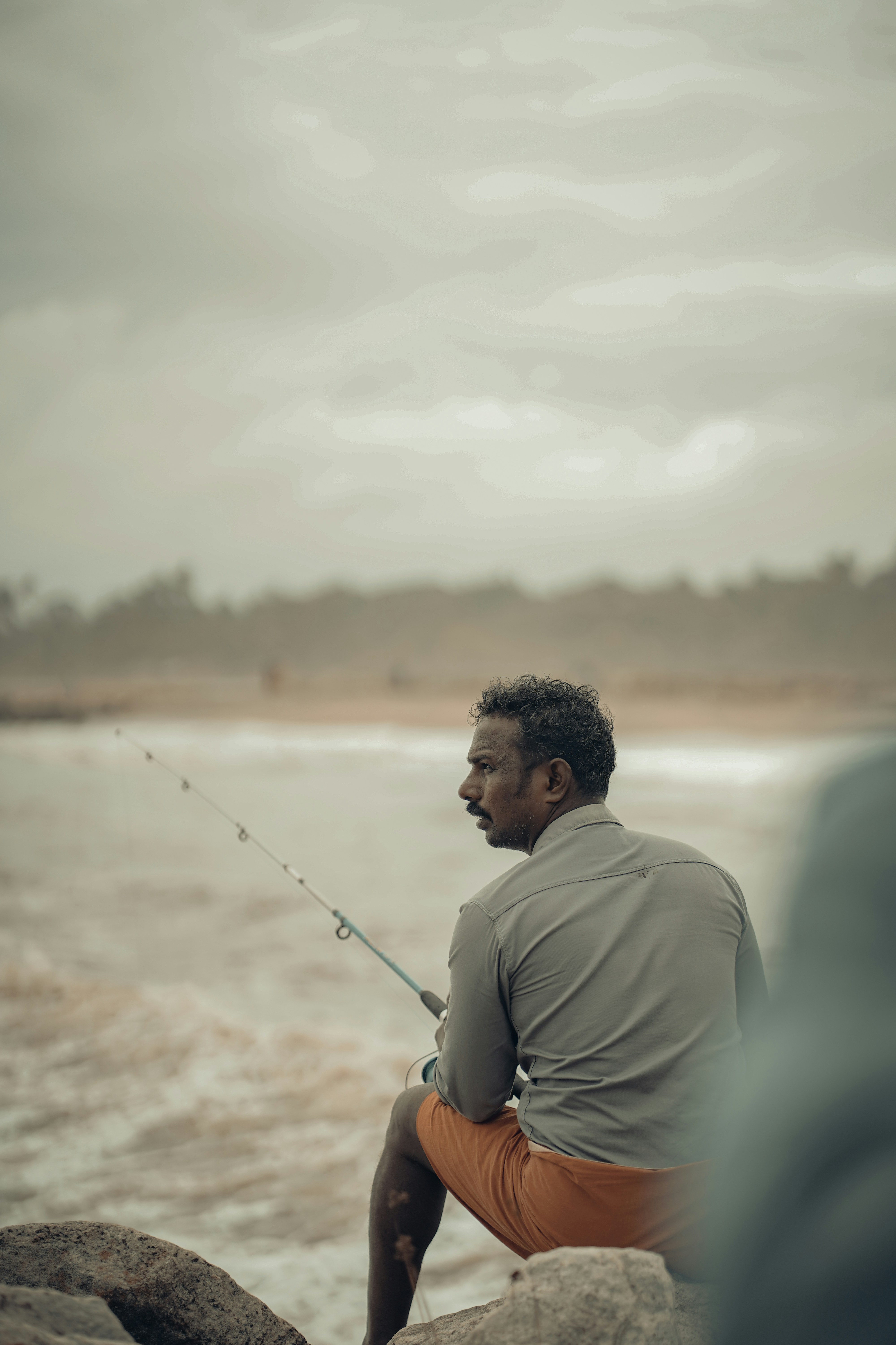 Fisherman seated on rocks, casting his line into turbulent waters under an overcast sky.