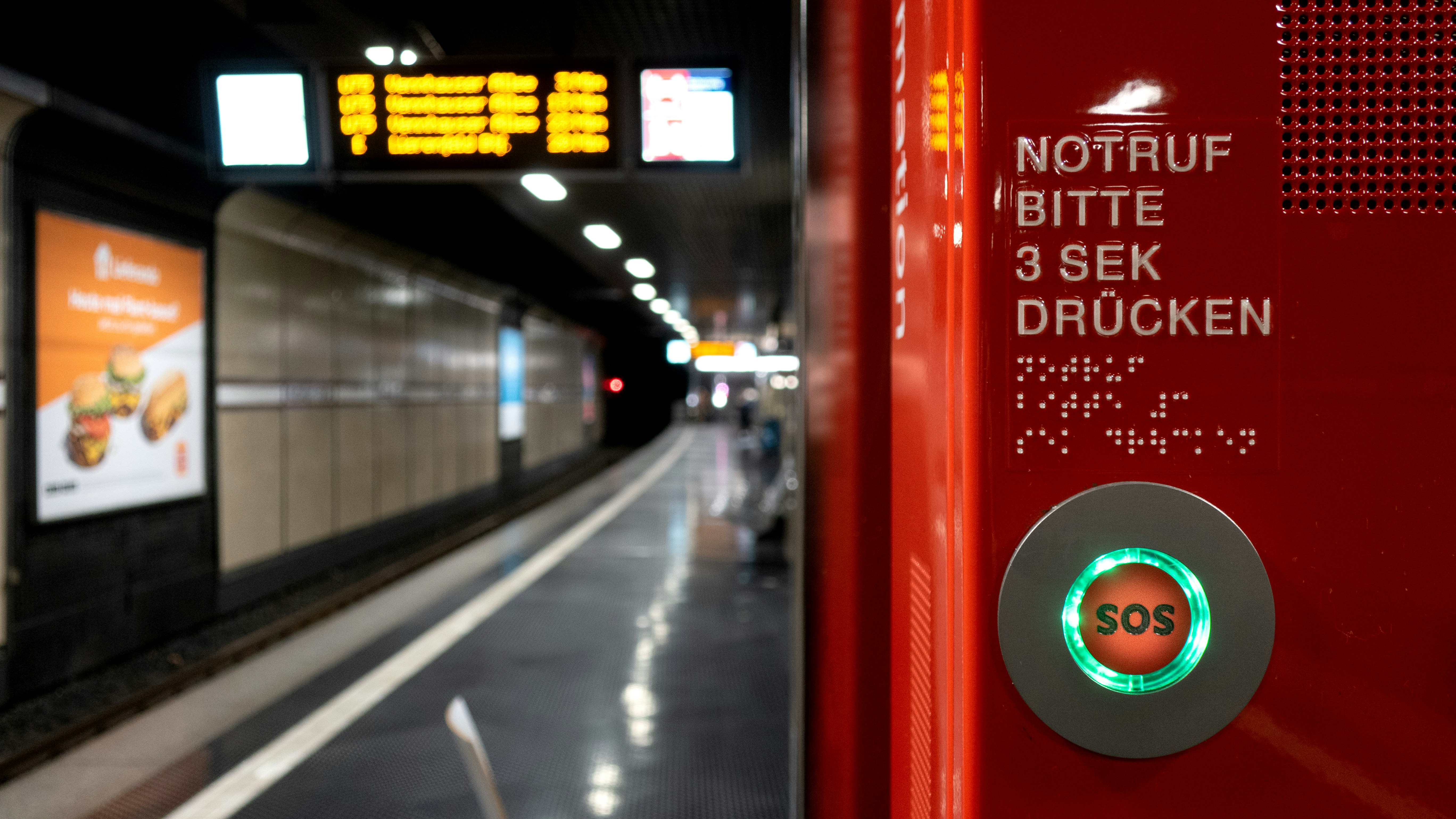 Red emergency button panel in a subway station with a blurred platform and signage in the background.