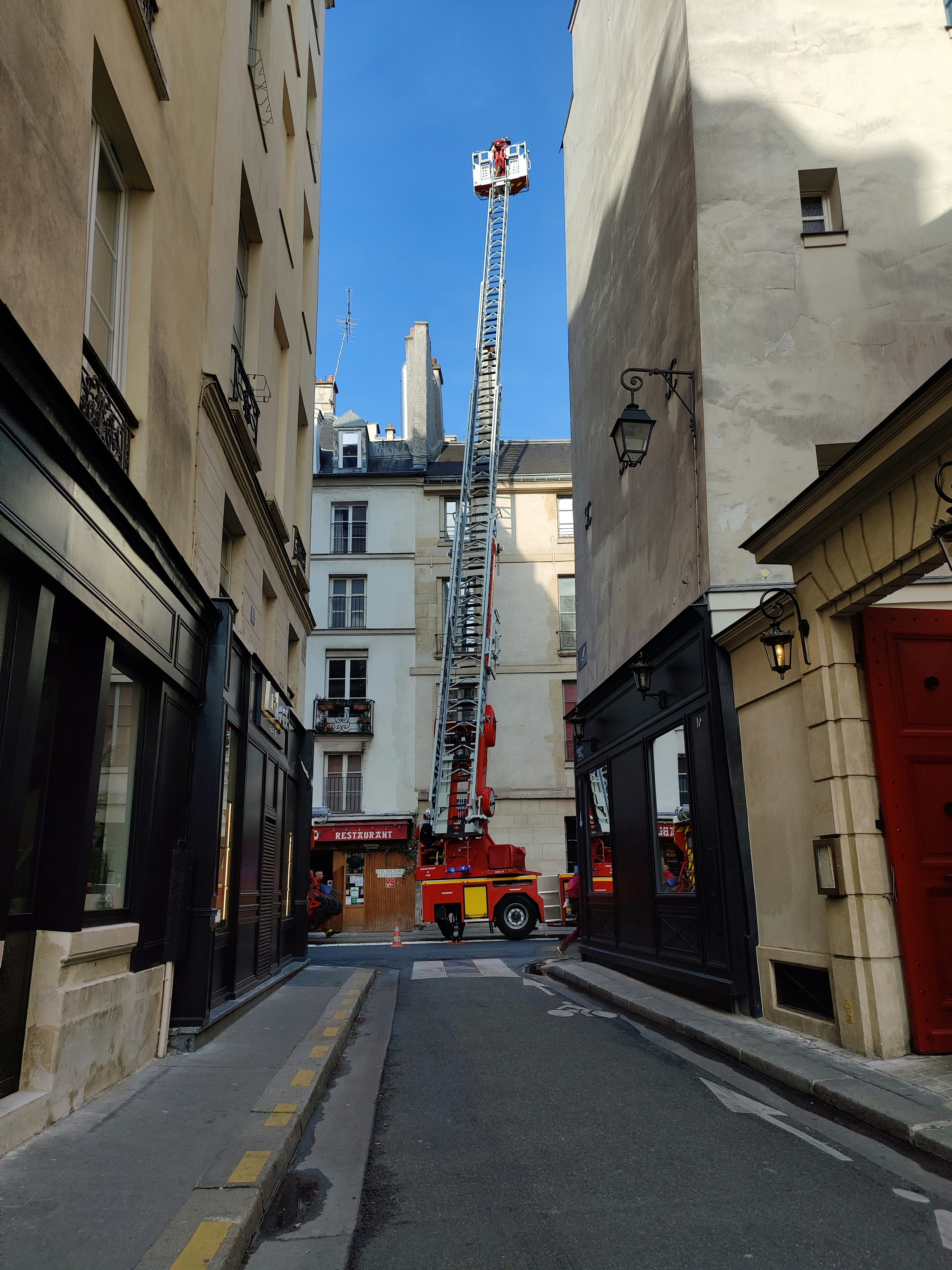 Photograph captures a red fire truck with an extended ladder reaching between tall beige buildings on a sunlit narrow street. The blue sky contrasts with the urban stonework.
