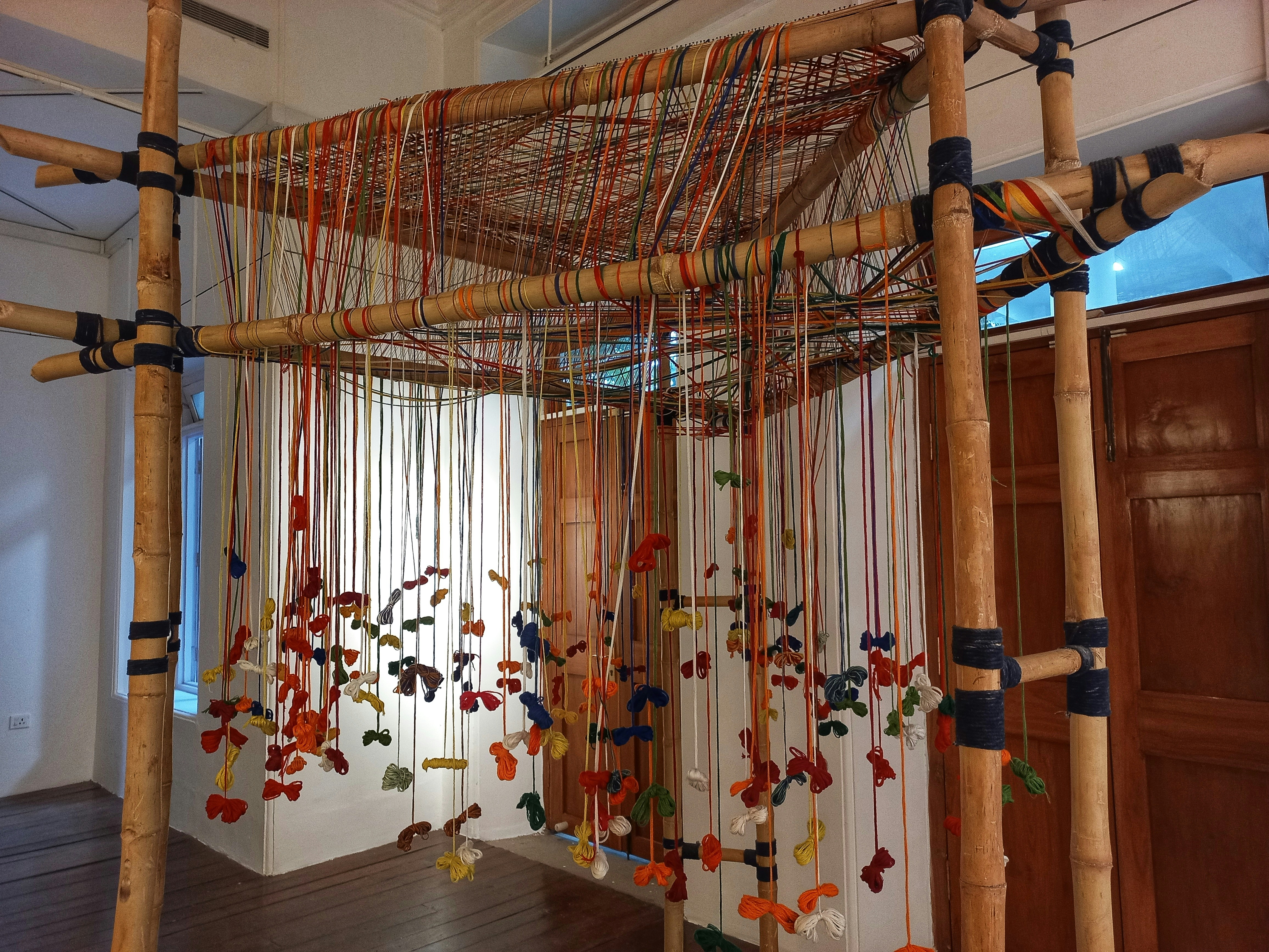 Colorful hanging threads weave through a wooden frame in an indoor installation, creating a playful curtain of hues and textures. A sculpture-like setup with rope and fabric elements fills the space.