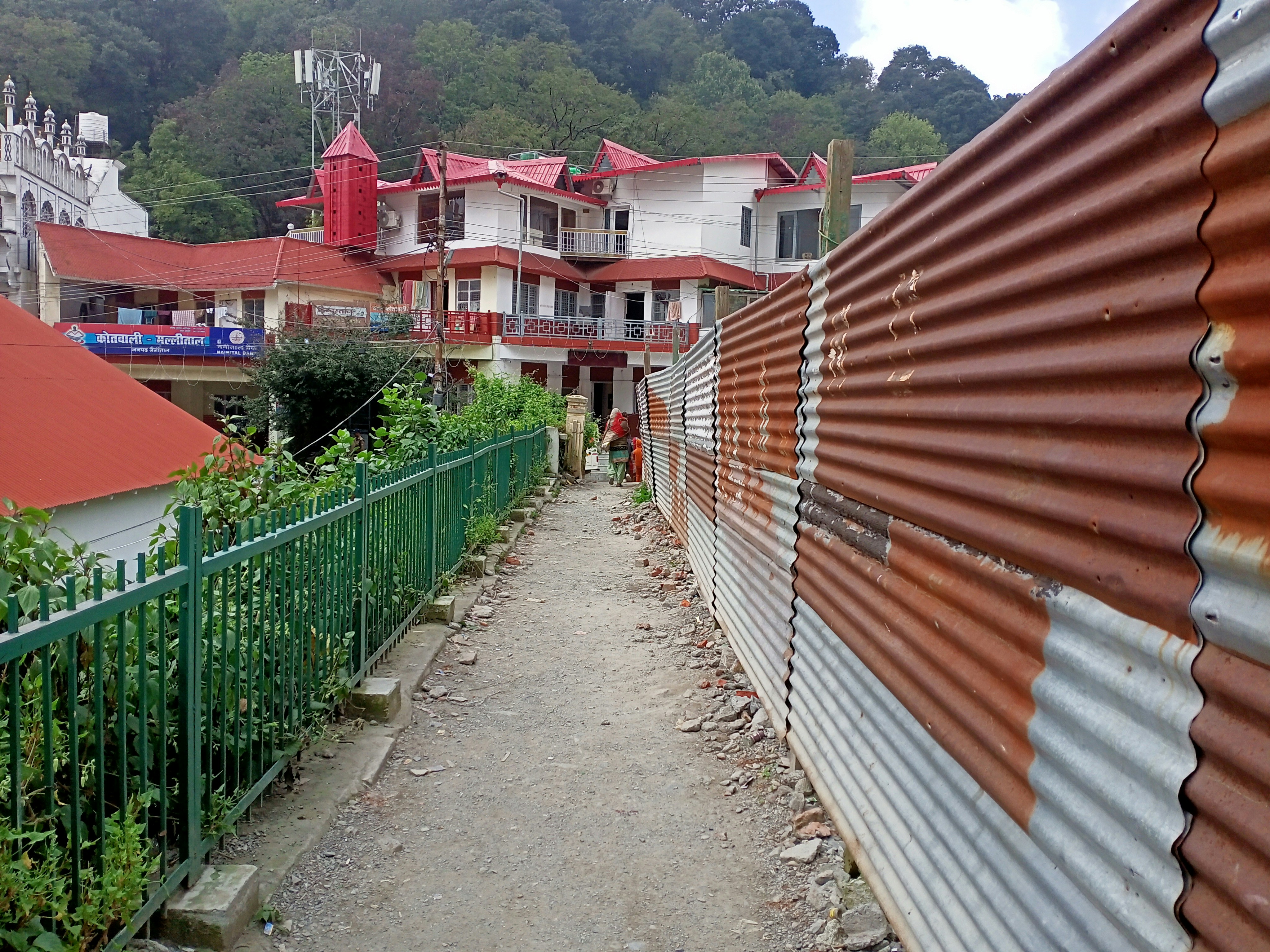 Narrow pathway lined with corrugated metal and greenery, leading to a cluster of colorful buildings in a hilly area.