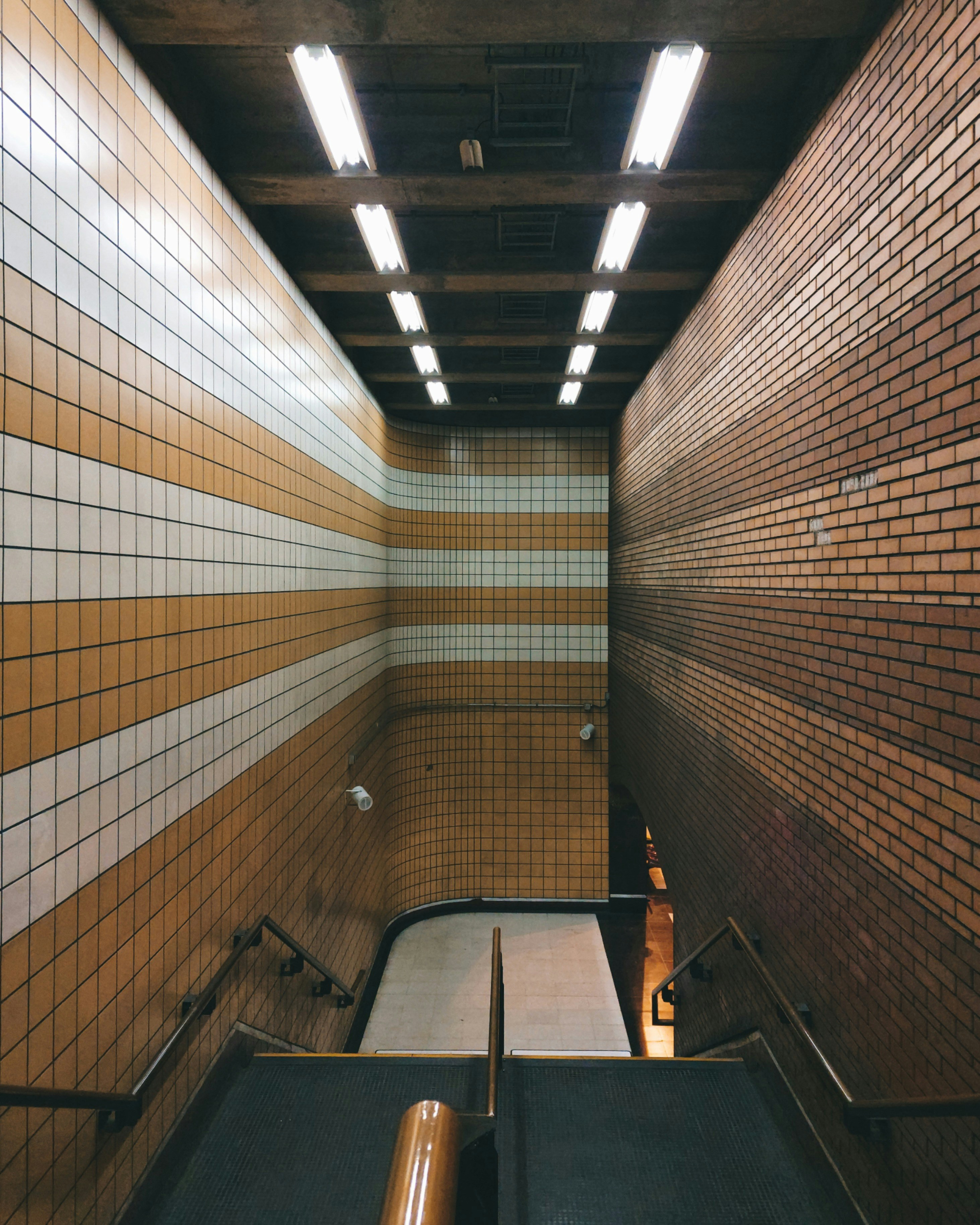 Staircase leading down into a tiled corridor with alternating stripes of color and light fixtures overhead.