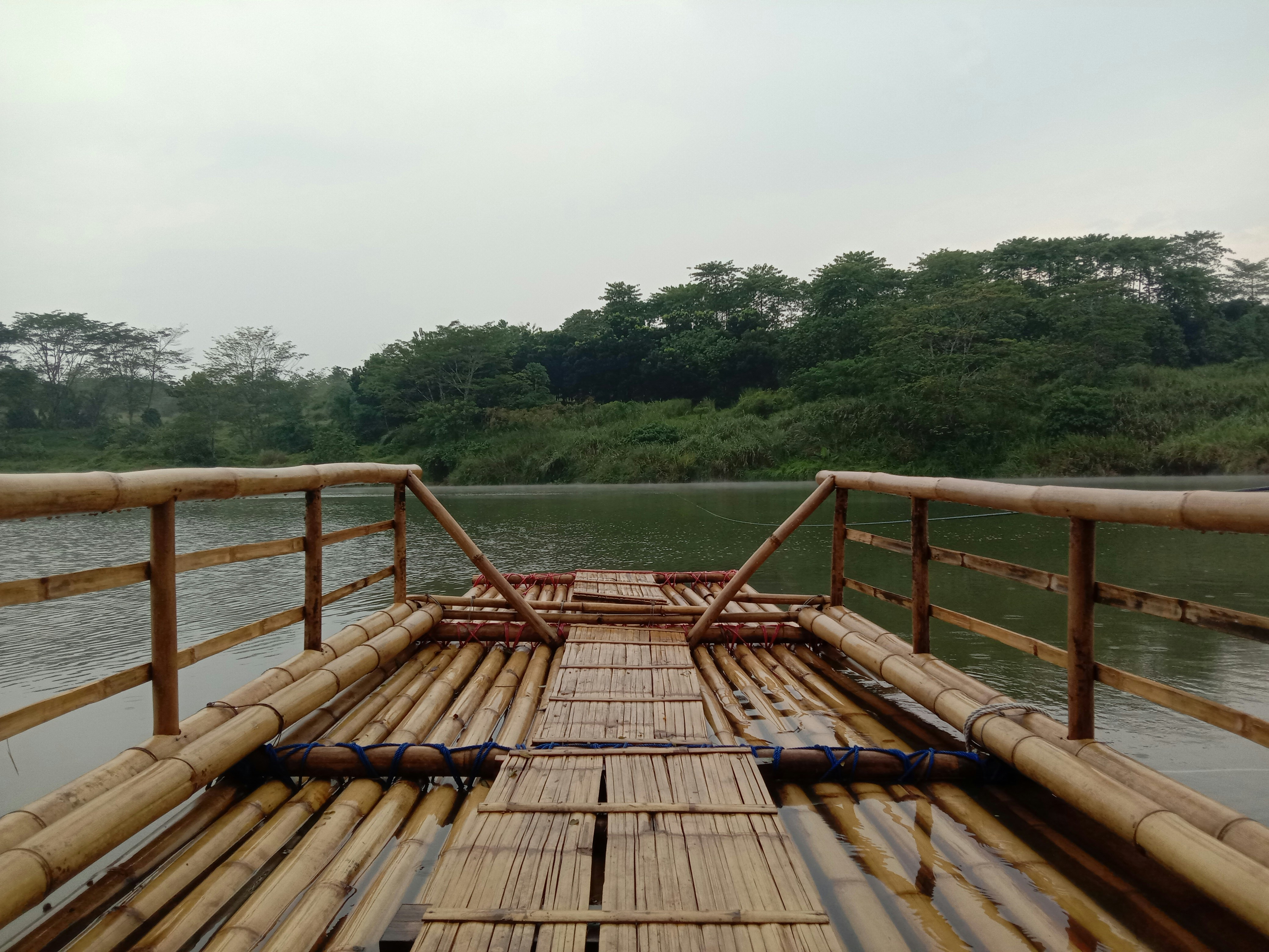Bamboo raft extending over a calm river, surrounded by lush greenery and a hazy sky. The scene invites tranquility and reflection.