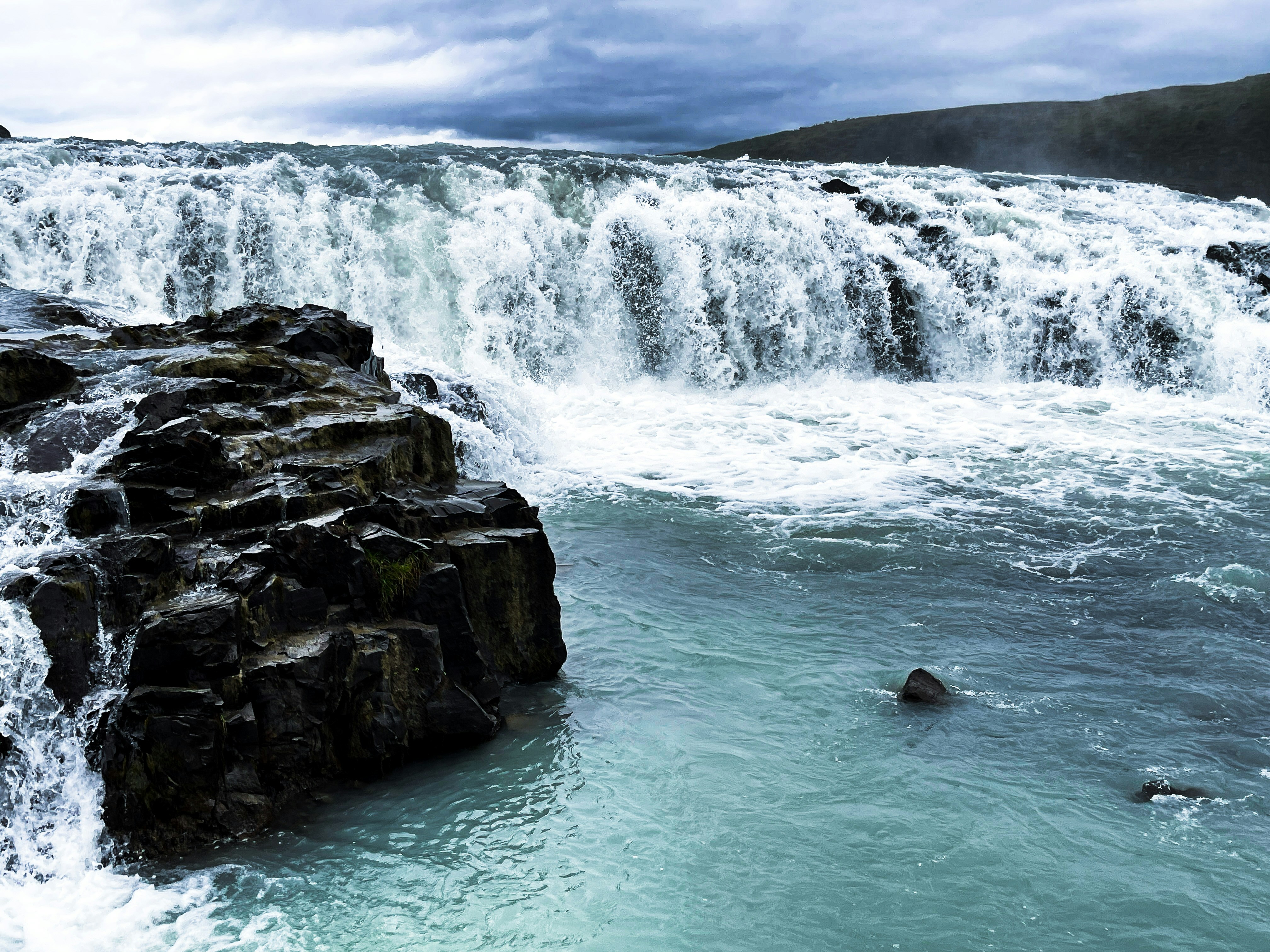 Powerful waterfall cascading over dark rocks into a turquoise pool, surrounded by a moody sky.
