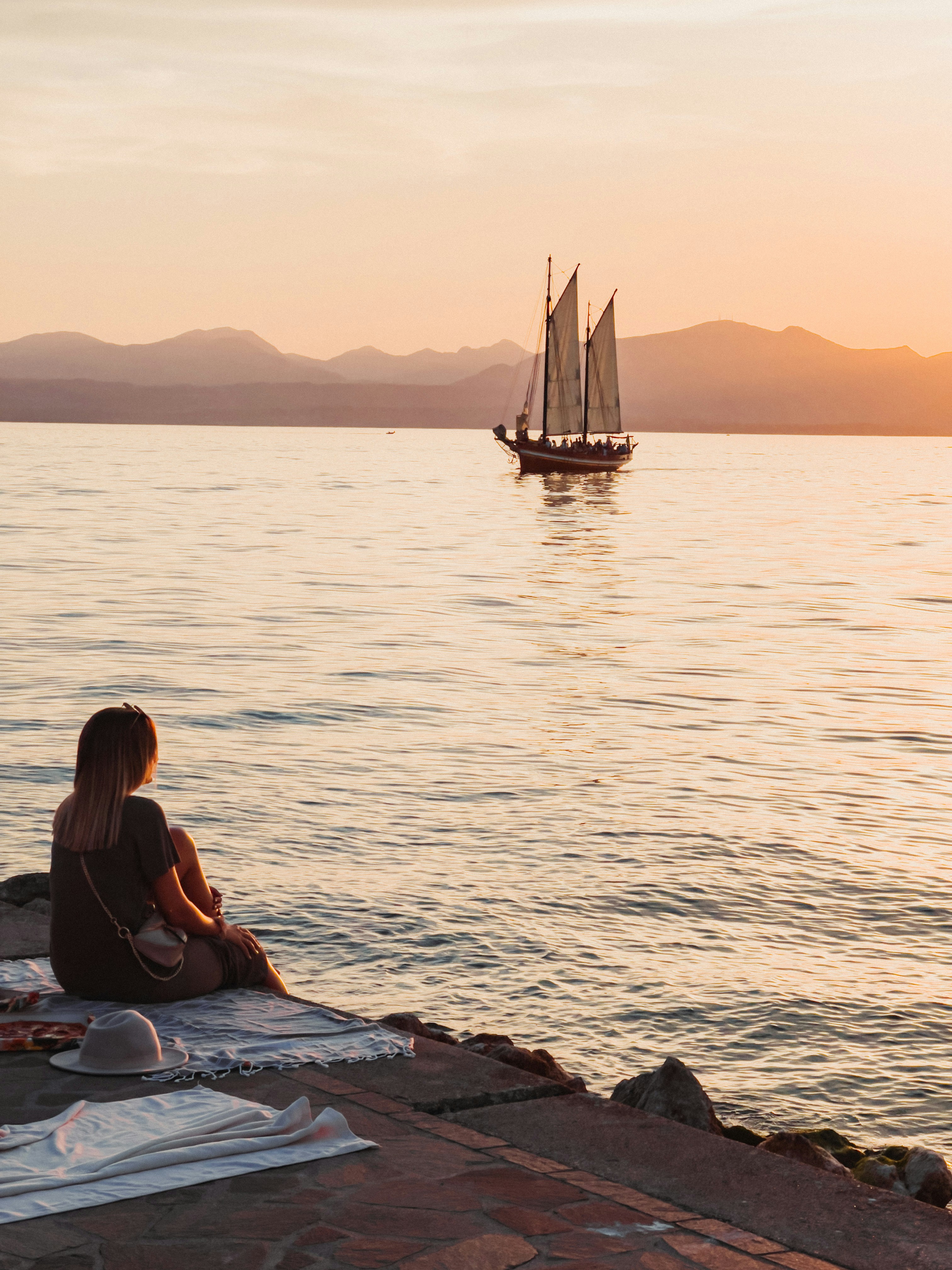 A sailboat glides across calm water at sunset as a person sits on a rocky jetty in the foreground.