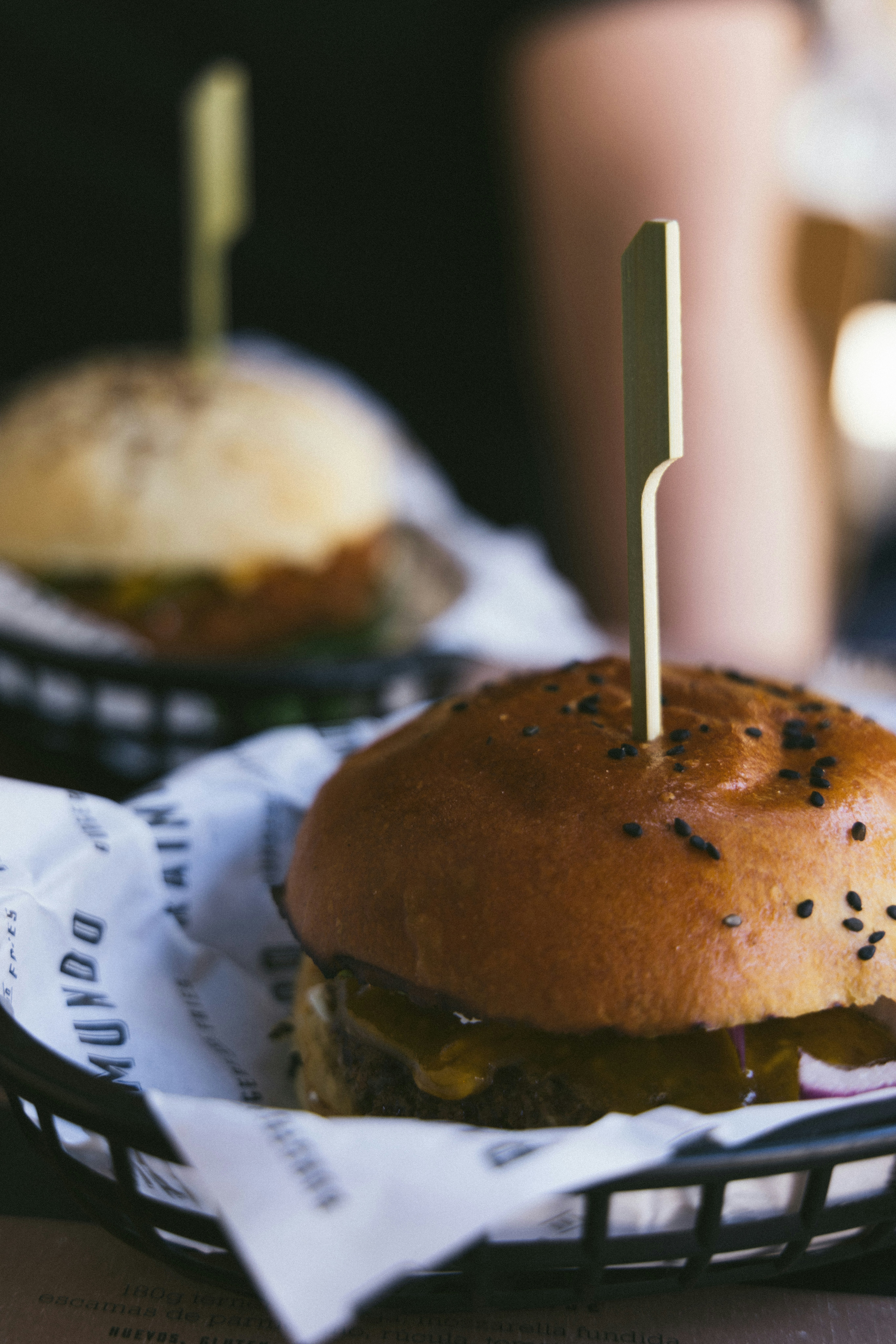 Gourmet burger topped with sesame seeds and a wooden skewer, nestled in a basket lined with parchment paper. A blurred background hints at a lively dining atmosphere.