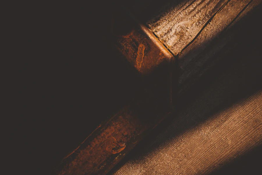 Close-up of a wooden surface or beam lit by warm, directional light against a dark background. The grain and worn edges create a moody, abstract composition.