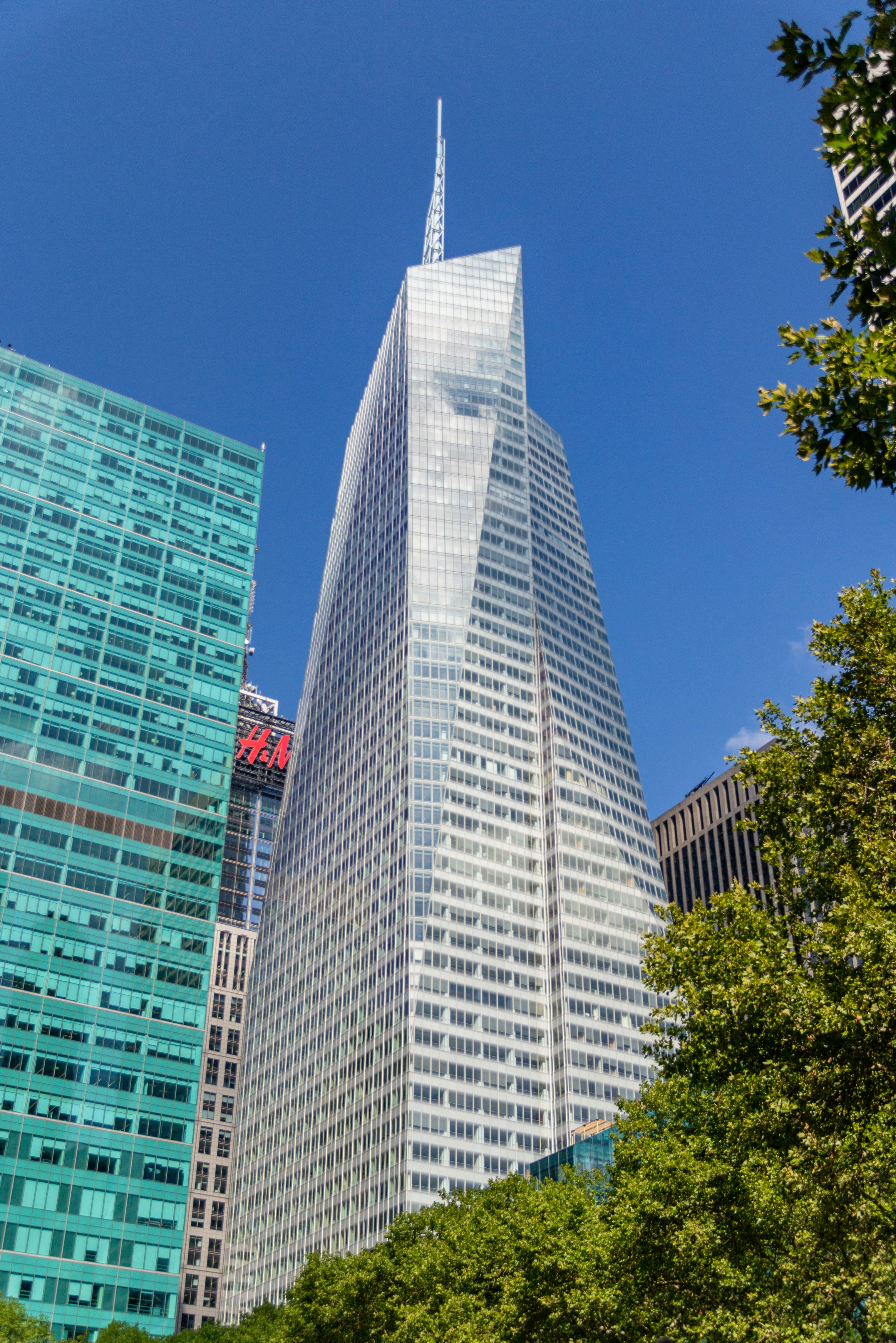 Sharp-edged skyscraper reflecting the sky, flanked by glass-clad buildings and lush greenery below.
