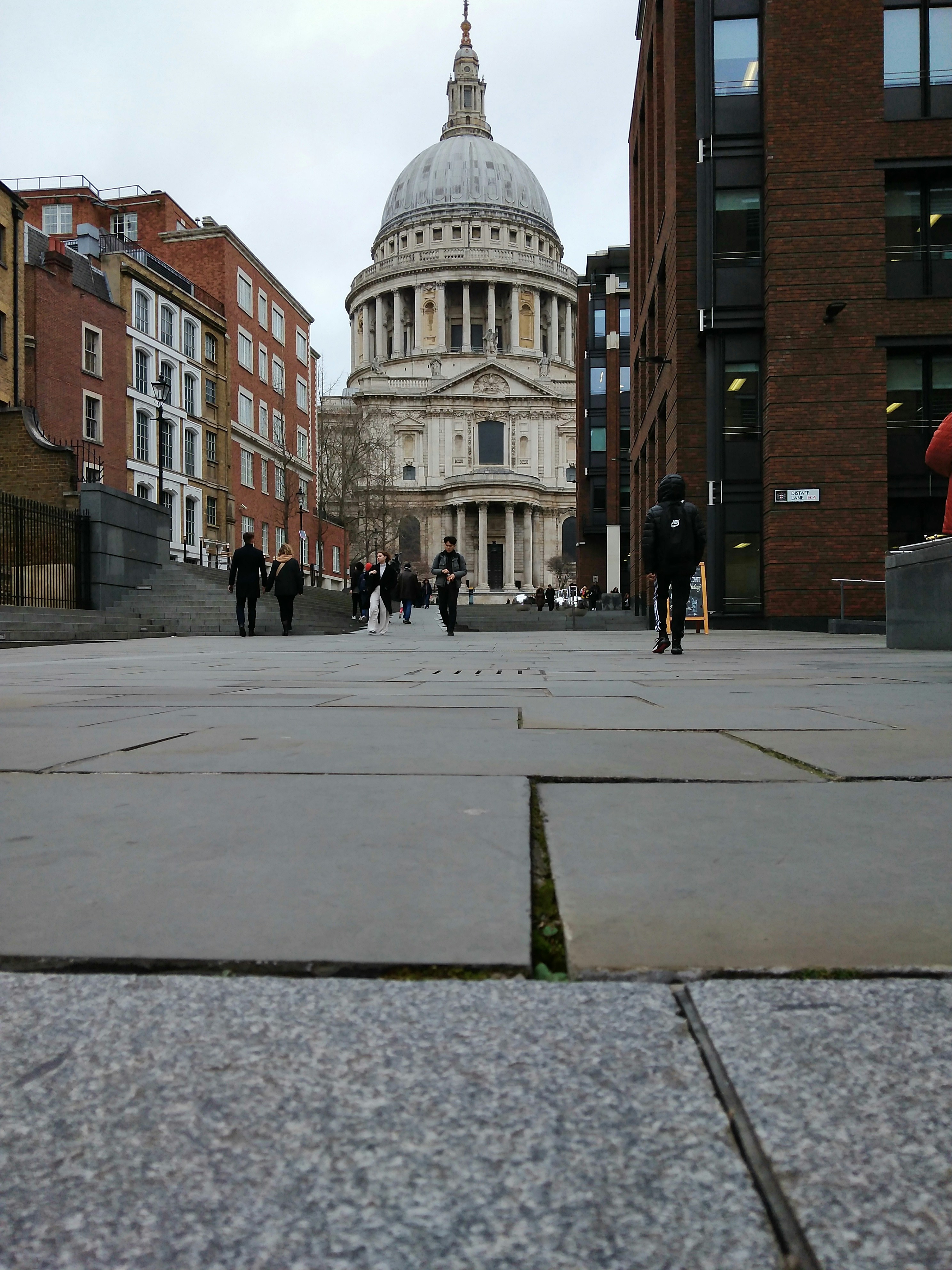 Wide-angle photograph of a city street leading toward St. Paul’s Cathedral, with pedestrians and brick facades framing the approach. Daylight capture emphasizes architectural detail and urban scale.