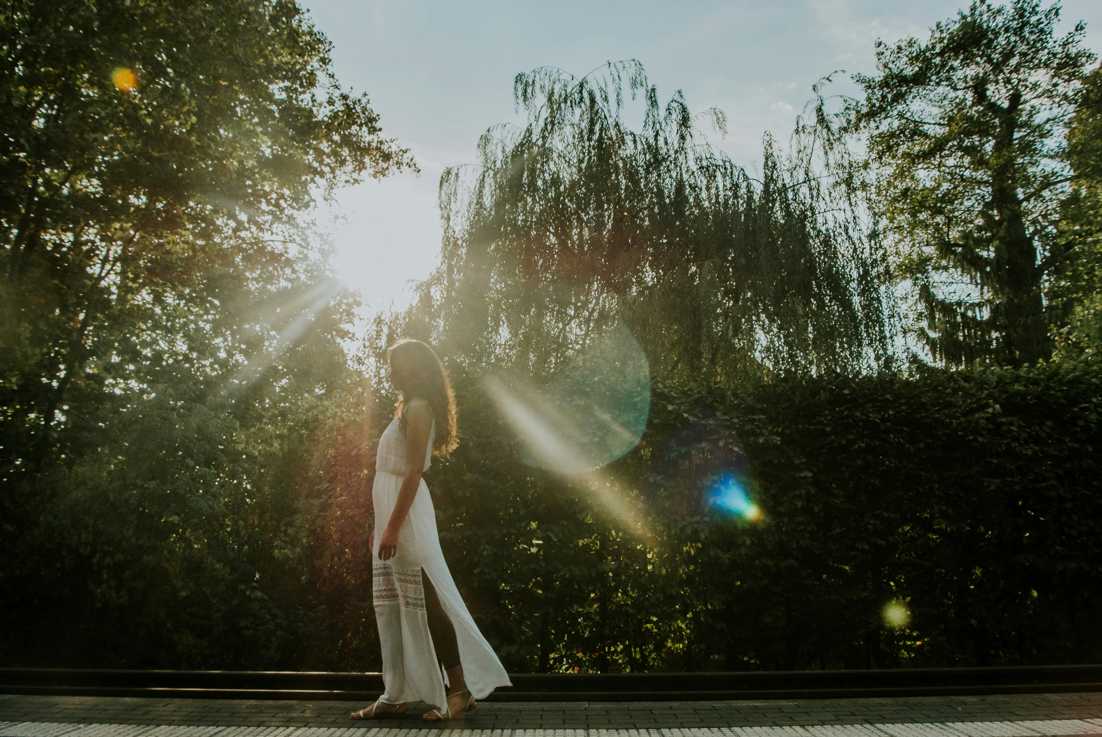 Woman in a white dress walking against the backdrop of sunlit trees, with lens flares adding a dreamy effect.