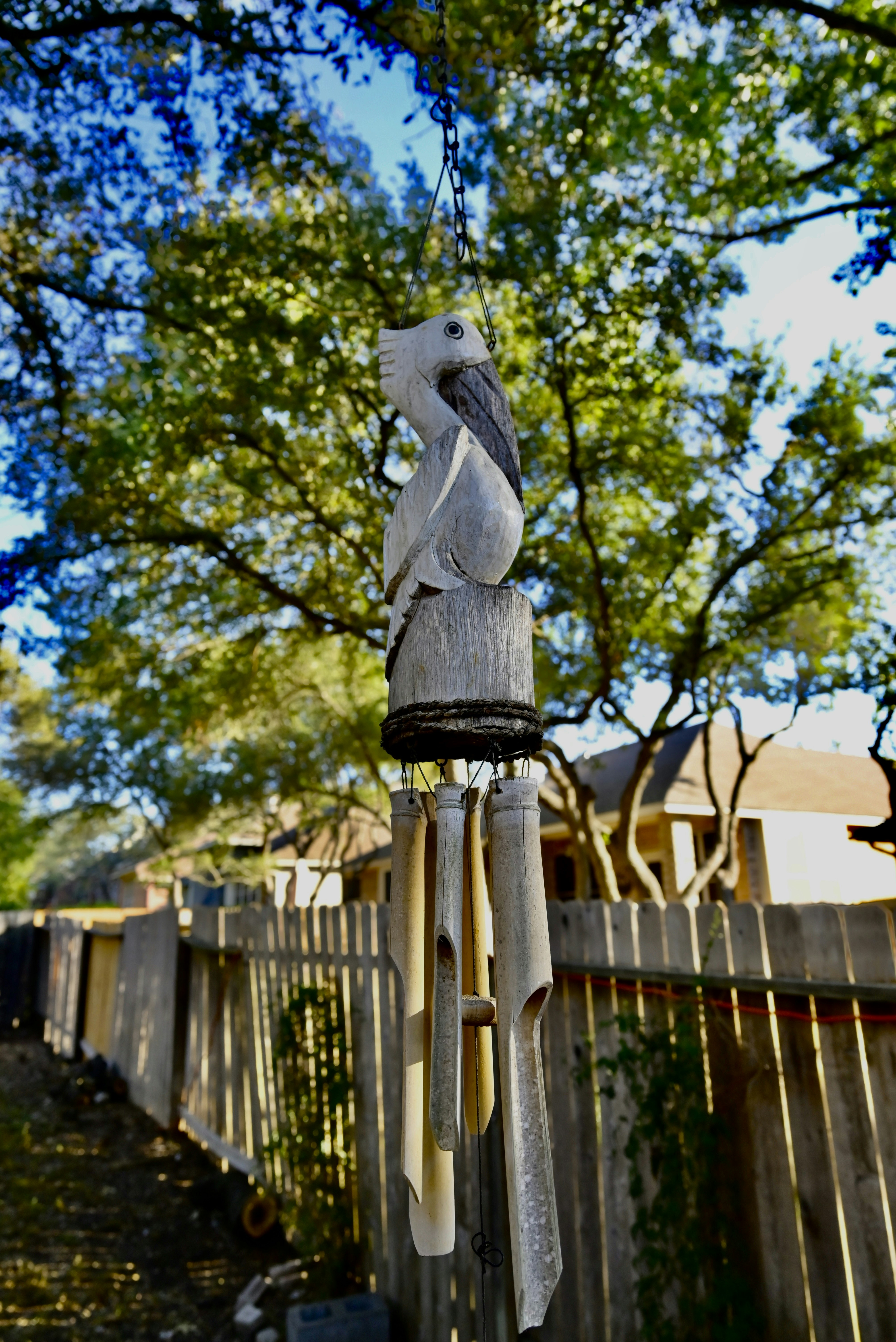 Handcrafted wooden wind chime featuring a bird perched atop, surrounded by lush greenery and a wooden fence. The scene captures a serene outdoor atmosphere.