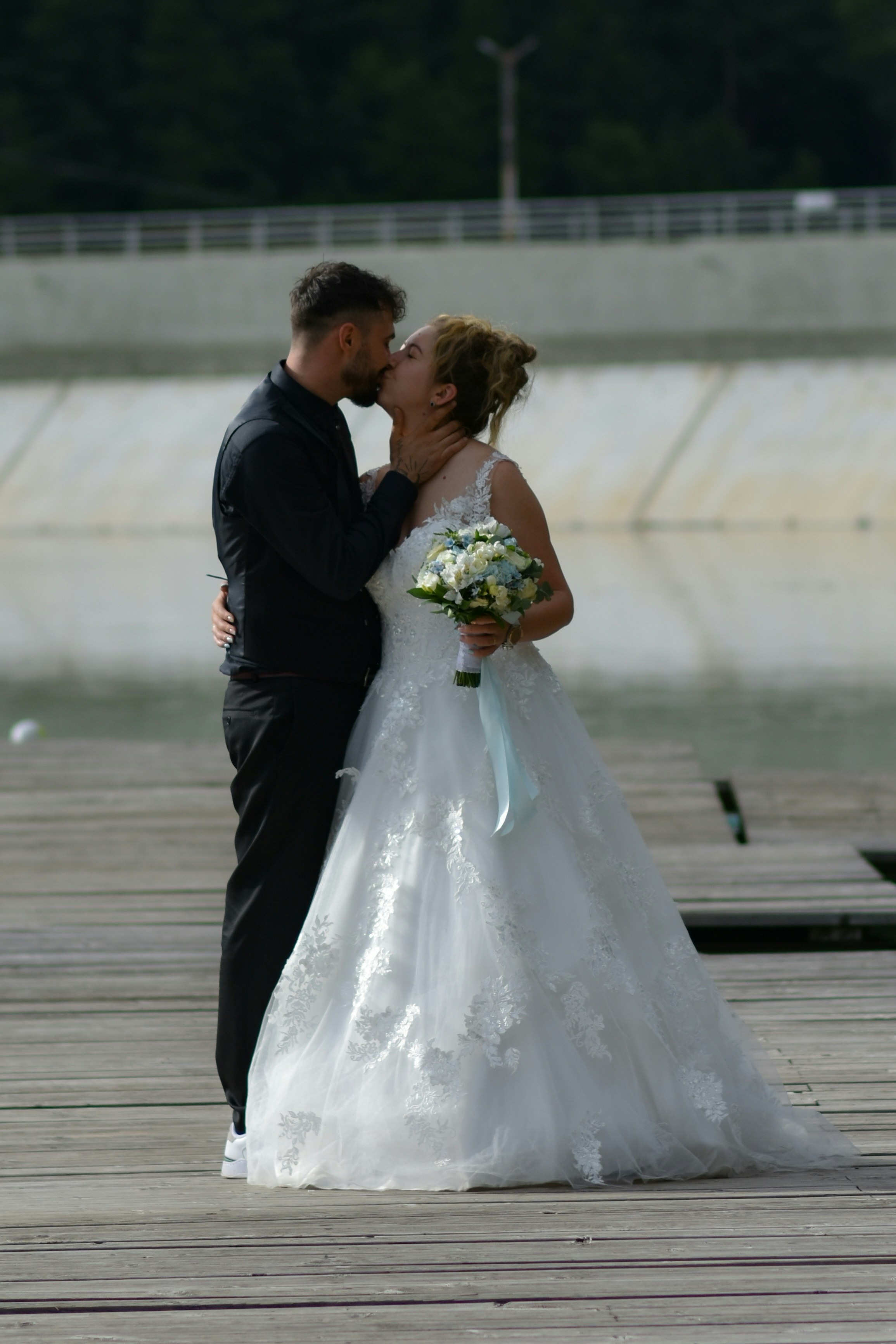 Bride and groom sharing a kiss on a wooden dock beside a still waterway.