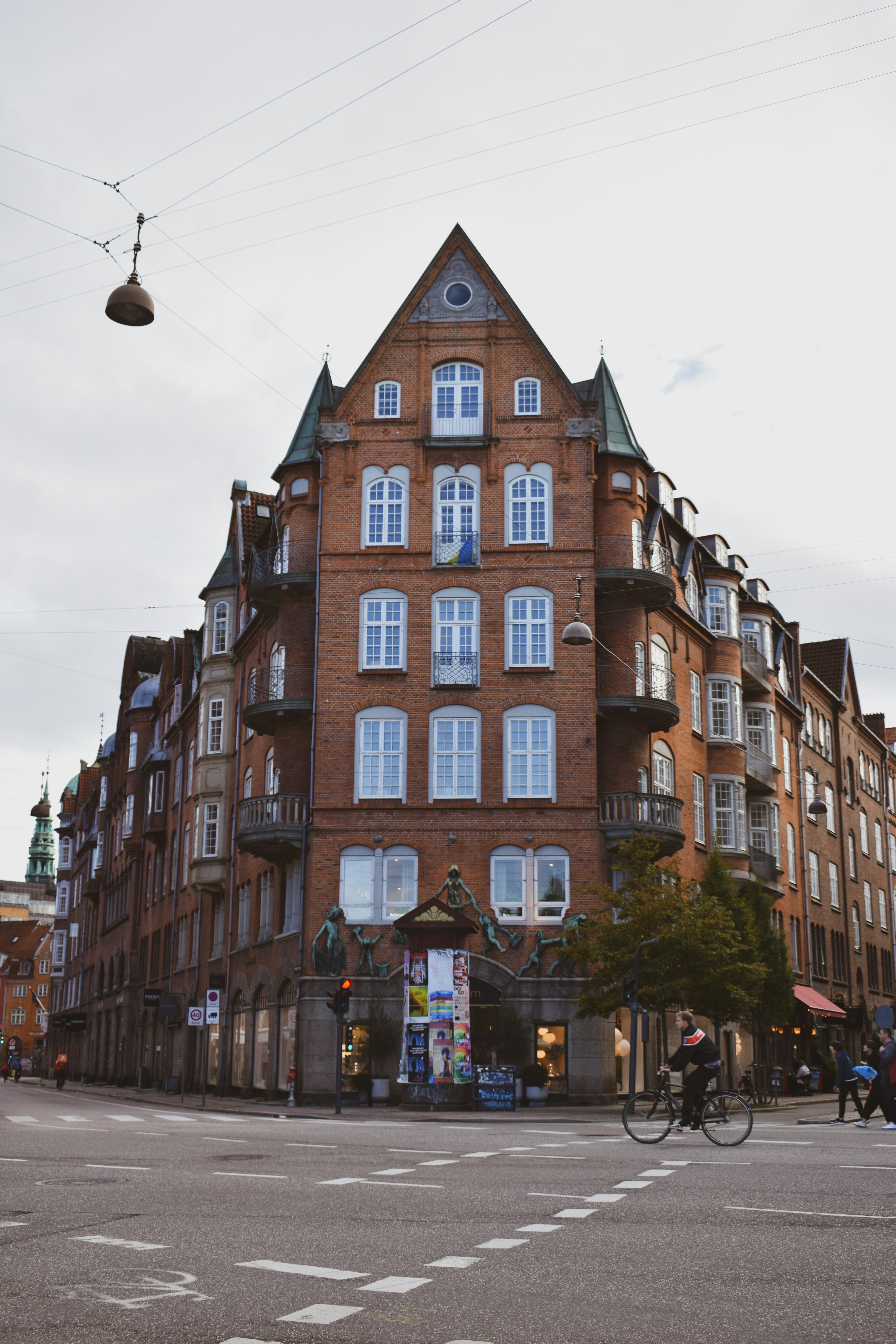 Historic brick building with intricate detailing and large windows, situated at a bustling street corner. A cyclist navigates the urban landscape.
