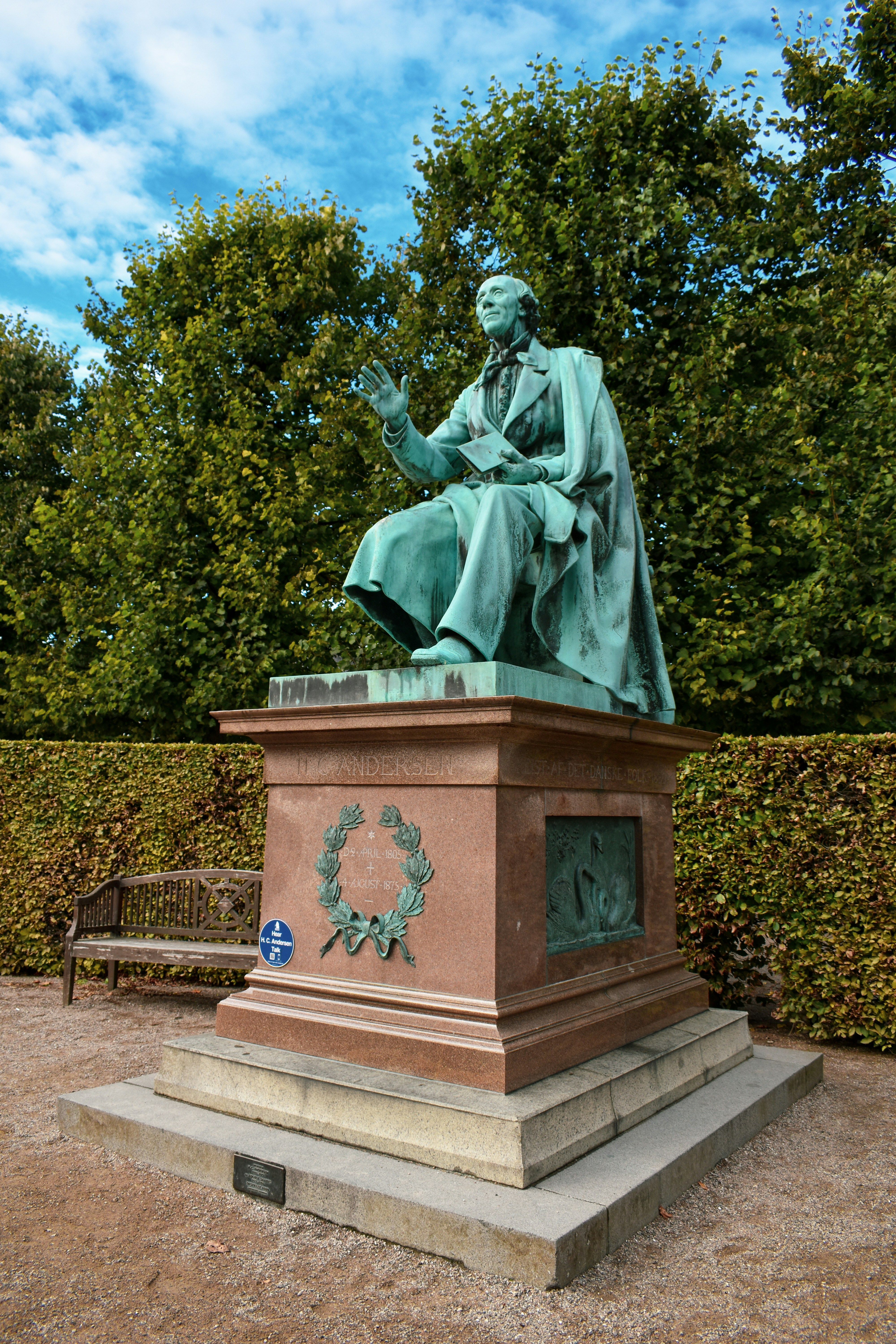 Teal patina bronze statue of a seated man with a raised hand on a red pedestal in a park, with a bench and trimmed hedges in the background.