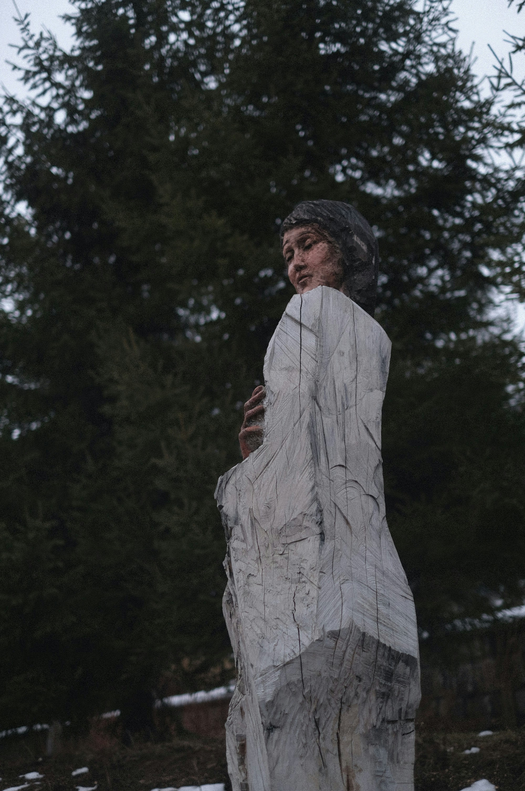 Intricately carved wooden statue of a woman standing amidst evergreen trees, exuding a serene presence. Snow blankets the ground, enhancing the tranquil atmosphere.