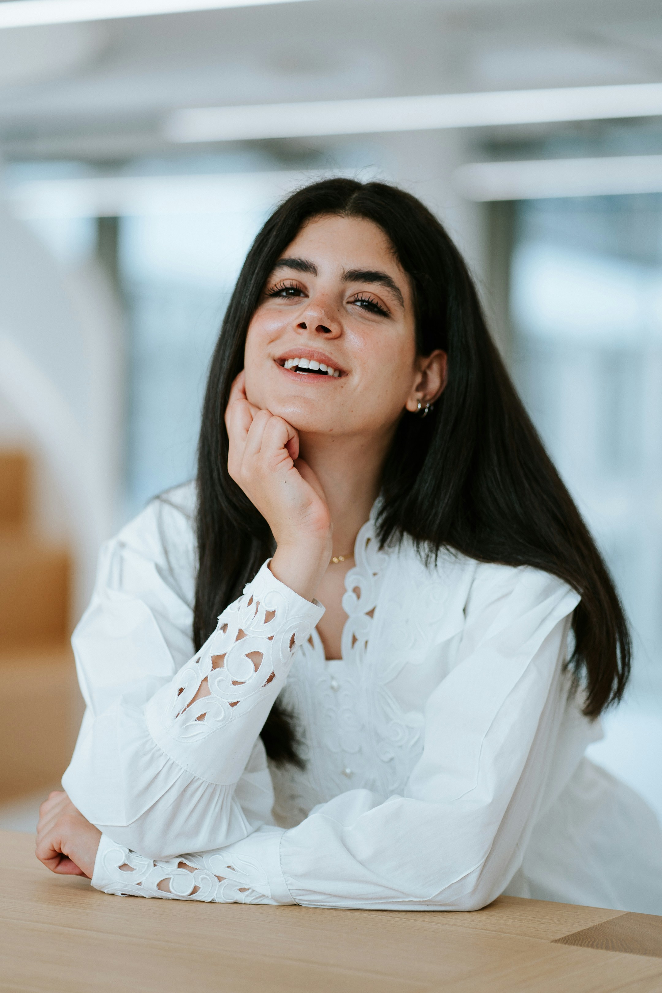 A woman with long black hair smiles thoughtfully while resting her chin on her hand, dressed in a white embroidered blouse. The background features a contemporary workspace.