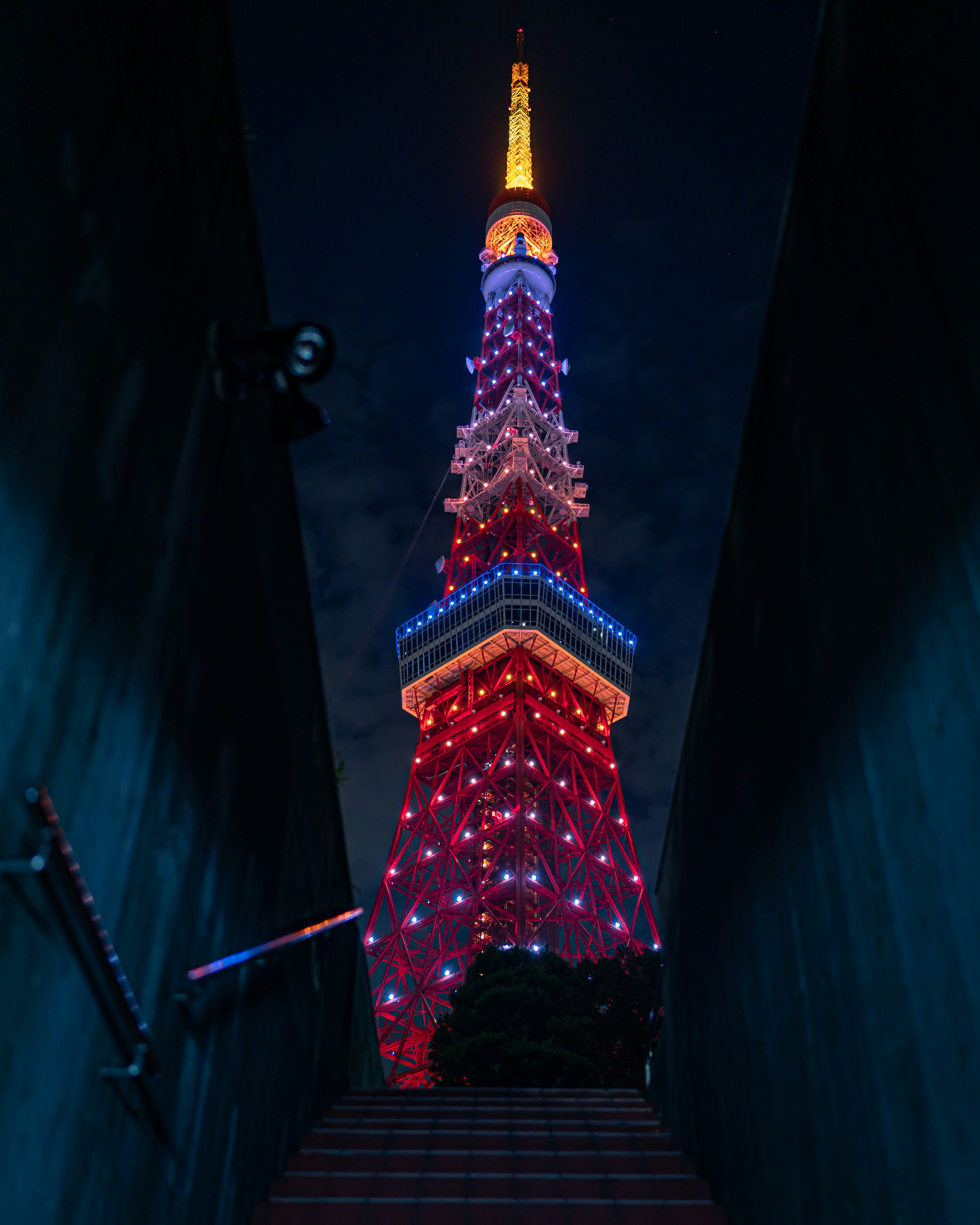 Tokyo Tower illuminated in vibrant red and blue lights, viewed from a stairway entrance at night.